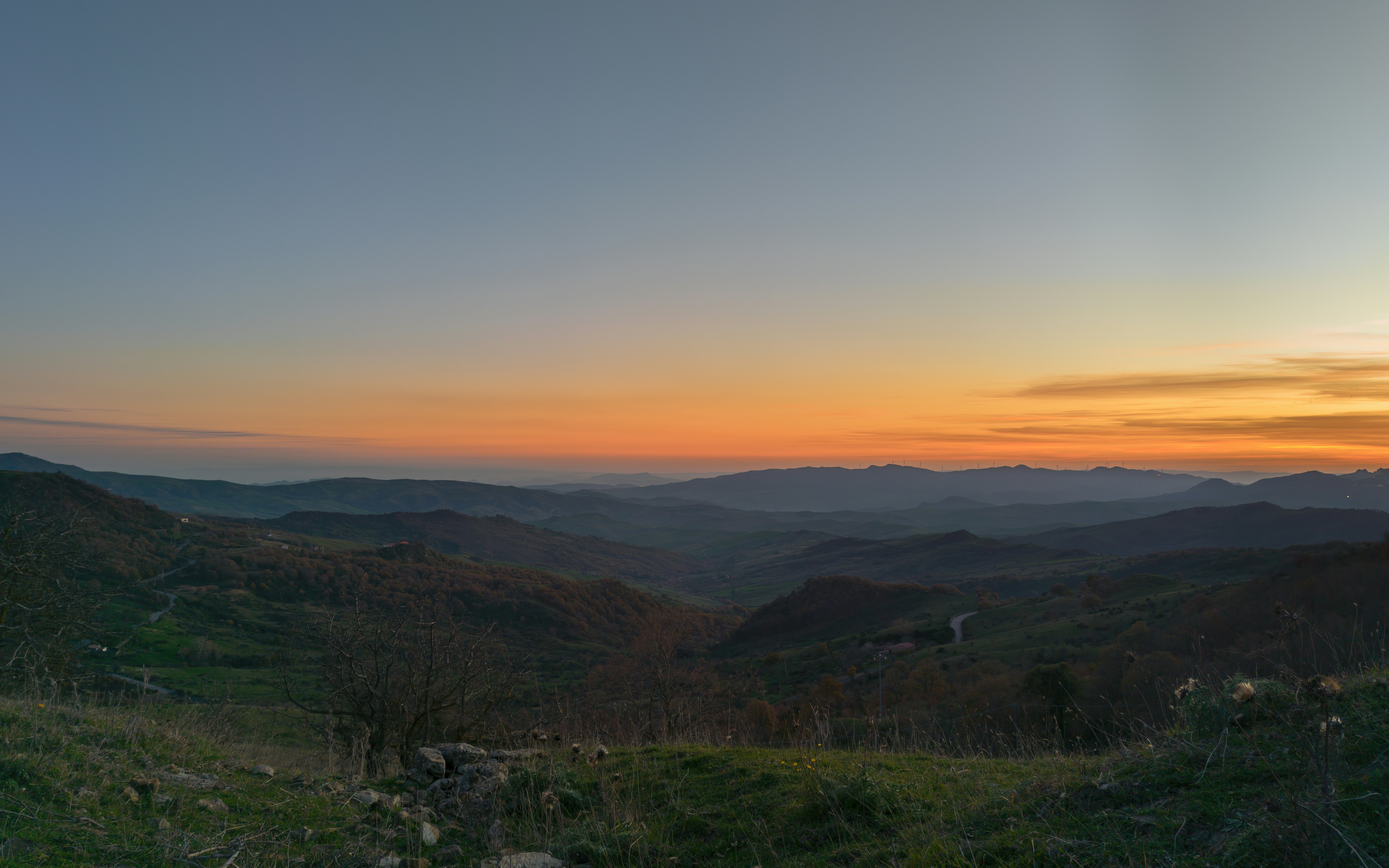 Sunset over rolling hills and distant ridges, with a warm orange horizon and a cool blue sky.