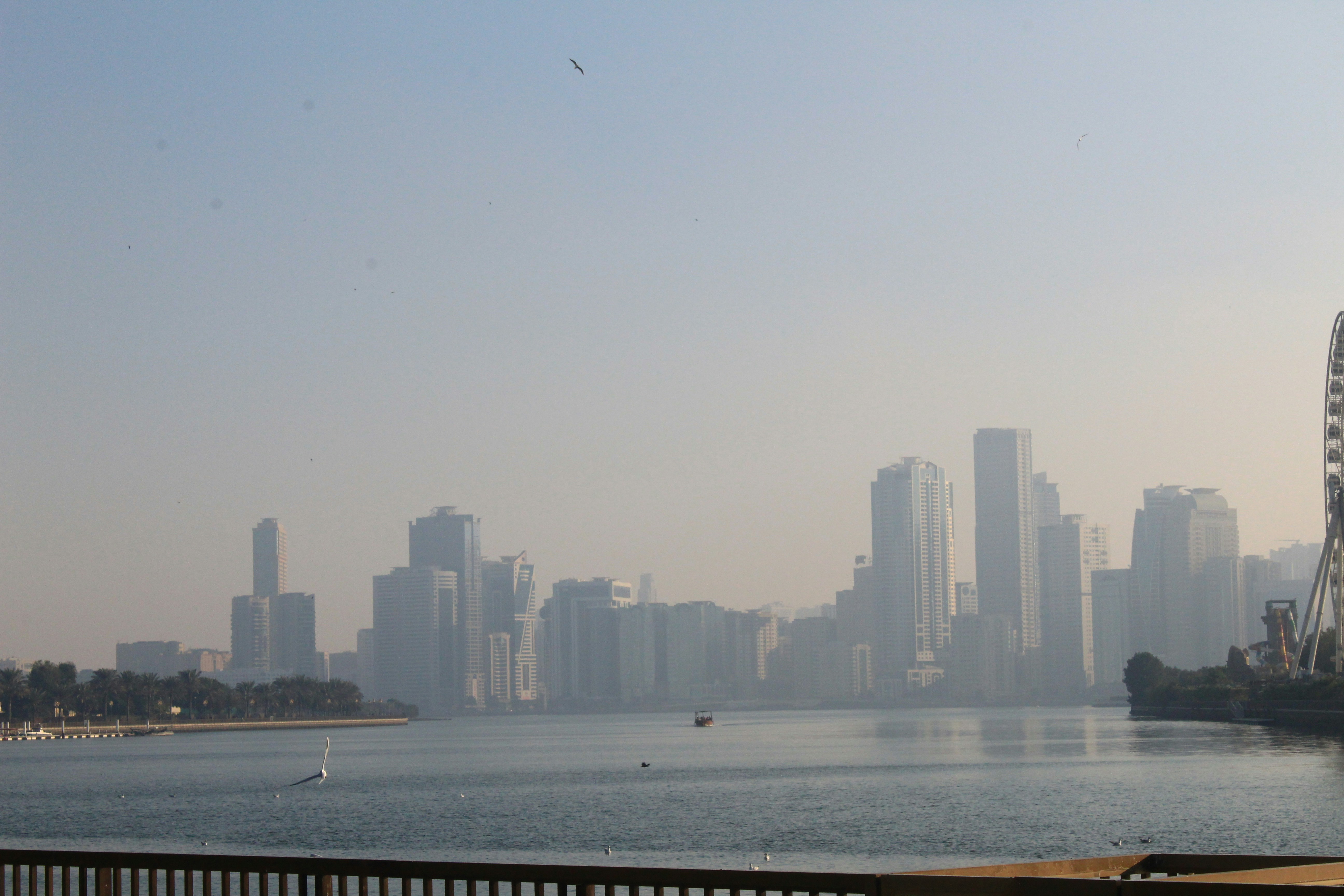 Urban skyline with tall buildings shrouded in mist, viewed across a calm body of water.