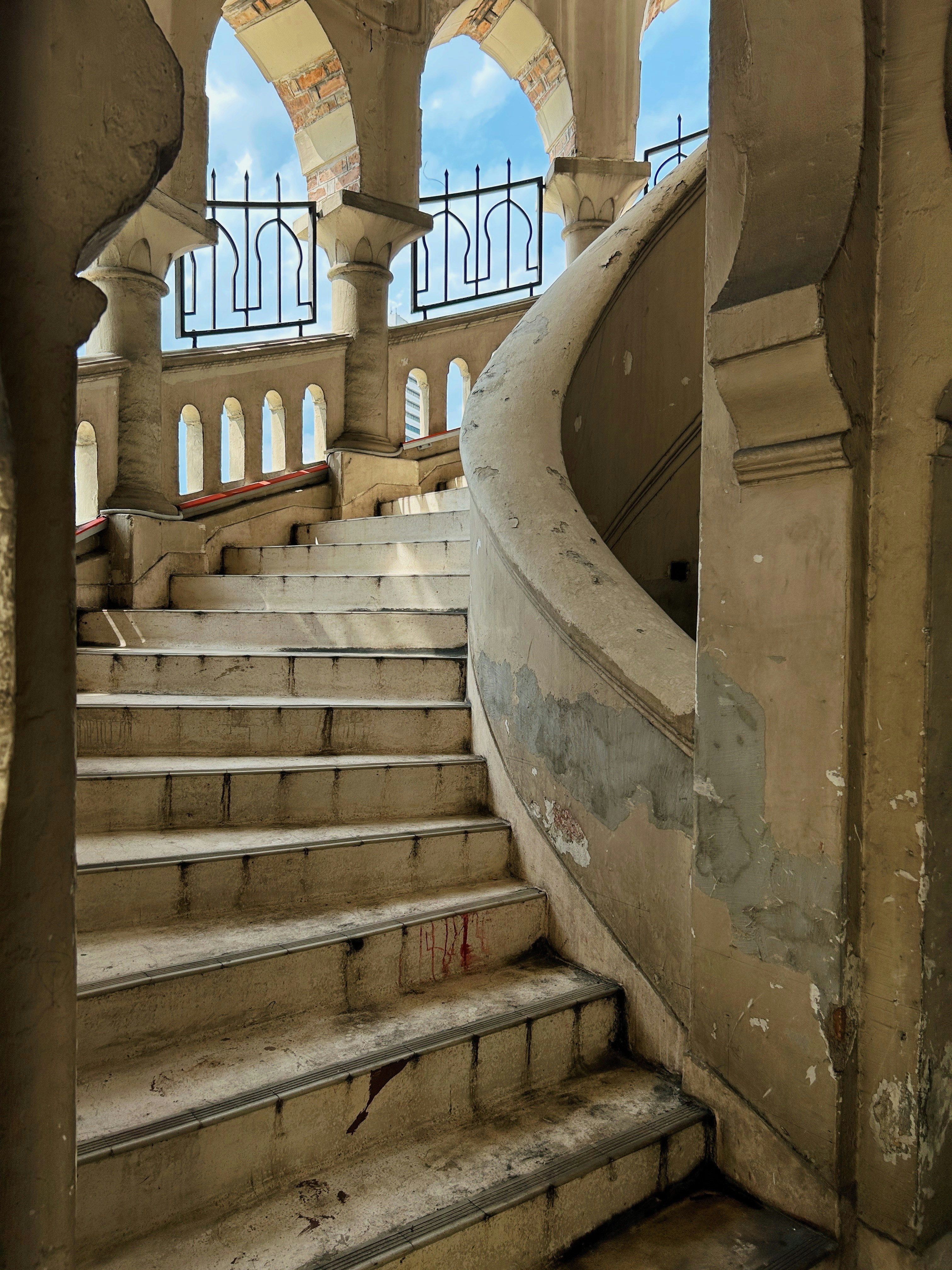 Curved staircase in an ornate, abandoned building, showcasing peeling paint and intricate architecture. Sunlight filters through arched windows, illuminating the steps.