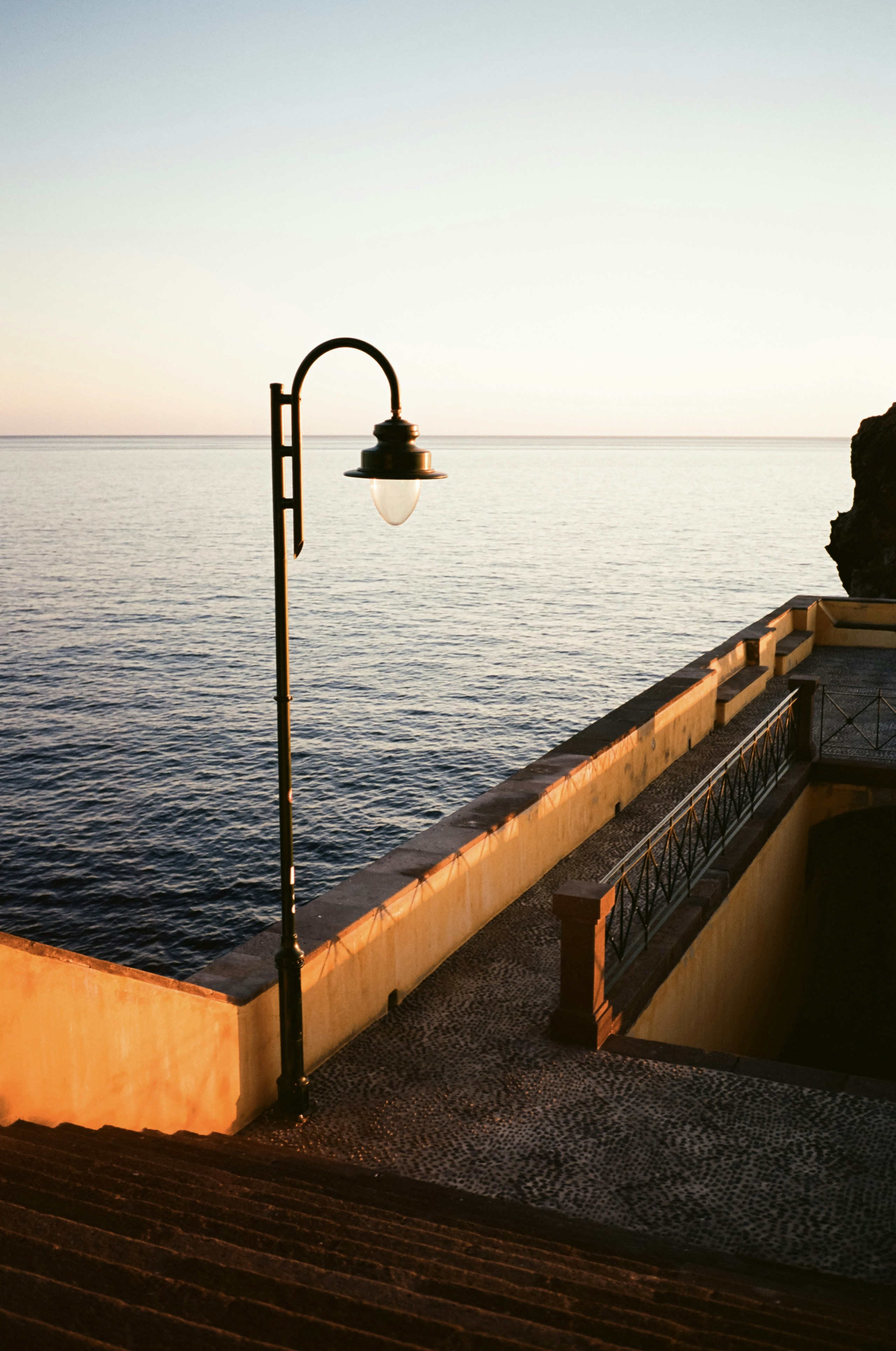 A vintage street lamp stands on a coastal walkway, casting a warm glow over the tranquil sea at dusk.