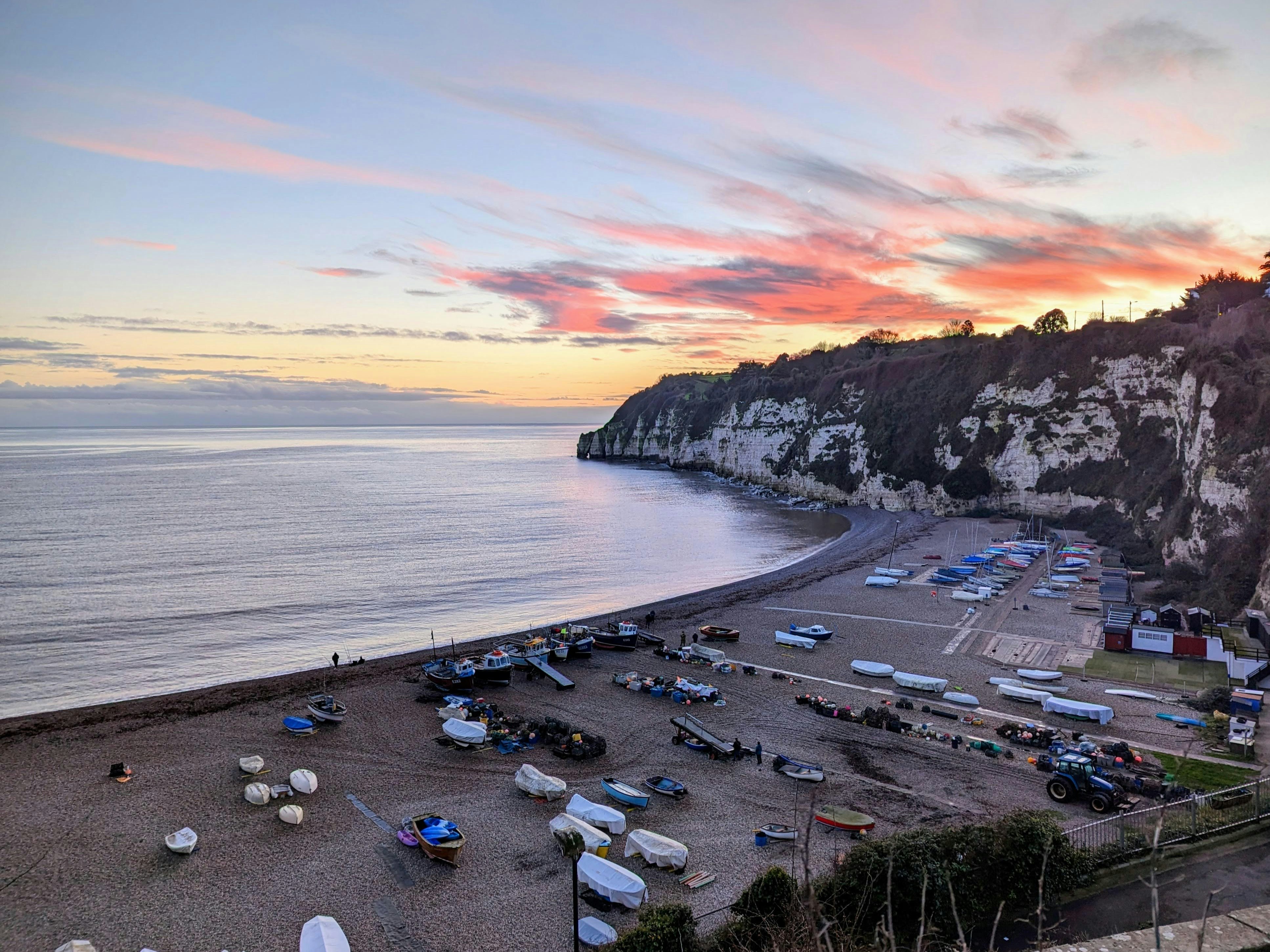 Coastal scene featuring a tranquil beach with boats resting on the shore, under a colorful sunset sky. The cliffs in the background add depth to the landscape.