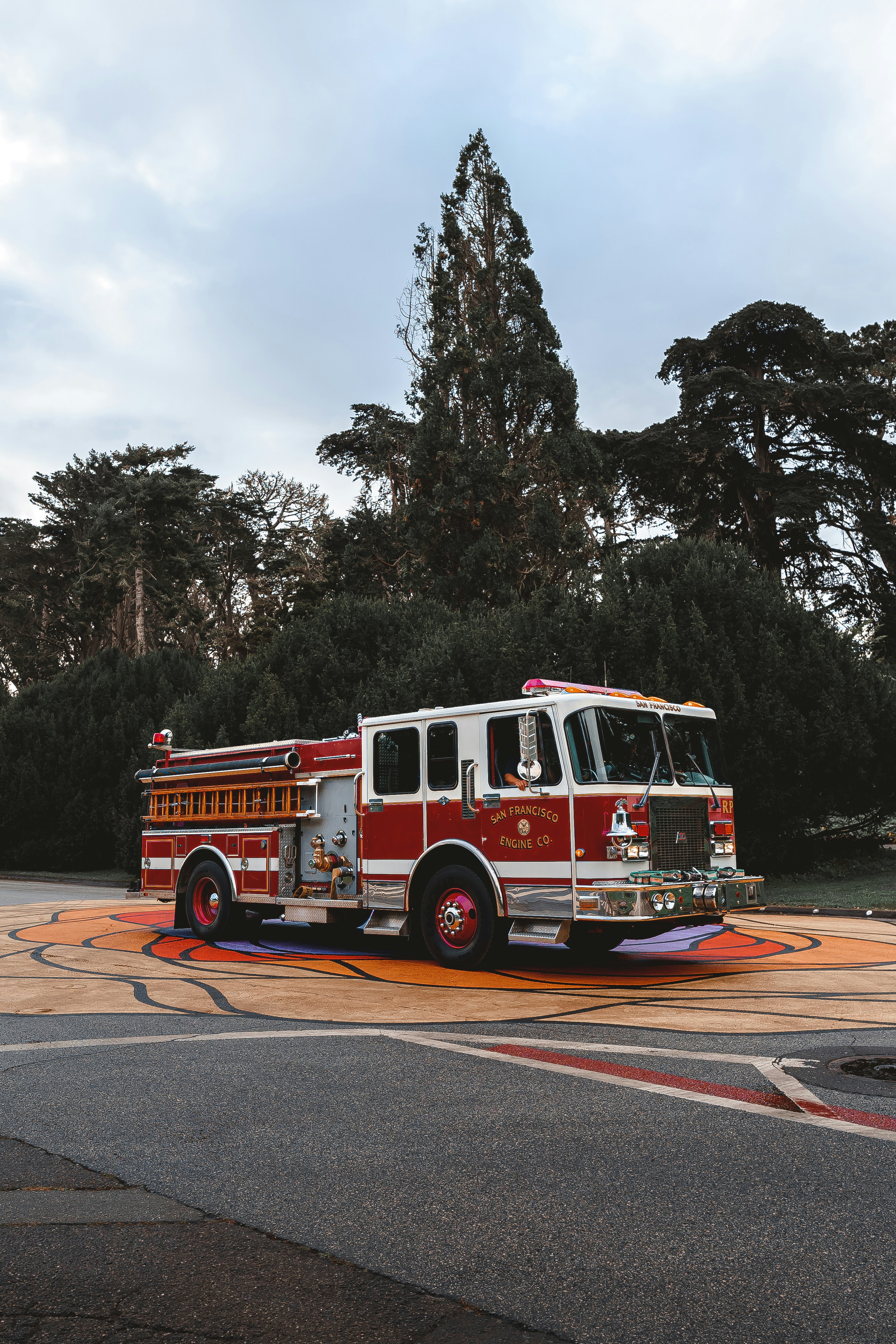A vibrant fire truck parked on a colorful street mural, surrounded by lush greenery. The scene captures the intersection of public service and artistic expression.