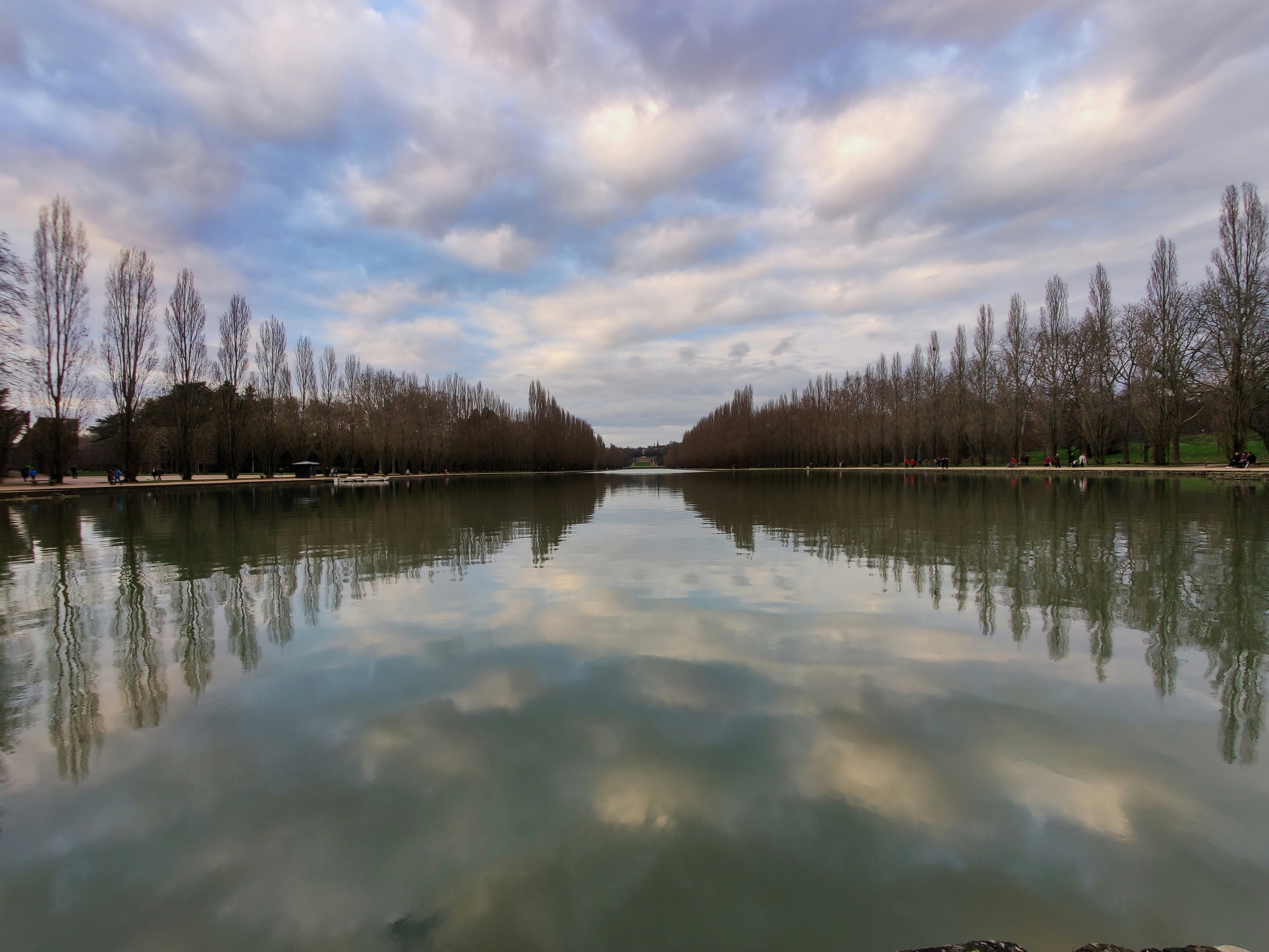 Calm canal flanked by leafless trees under a dramatic sky, with clouds mirrored in the water.
