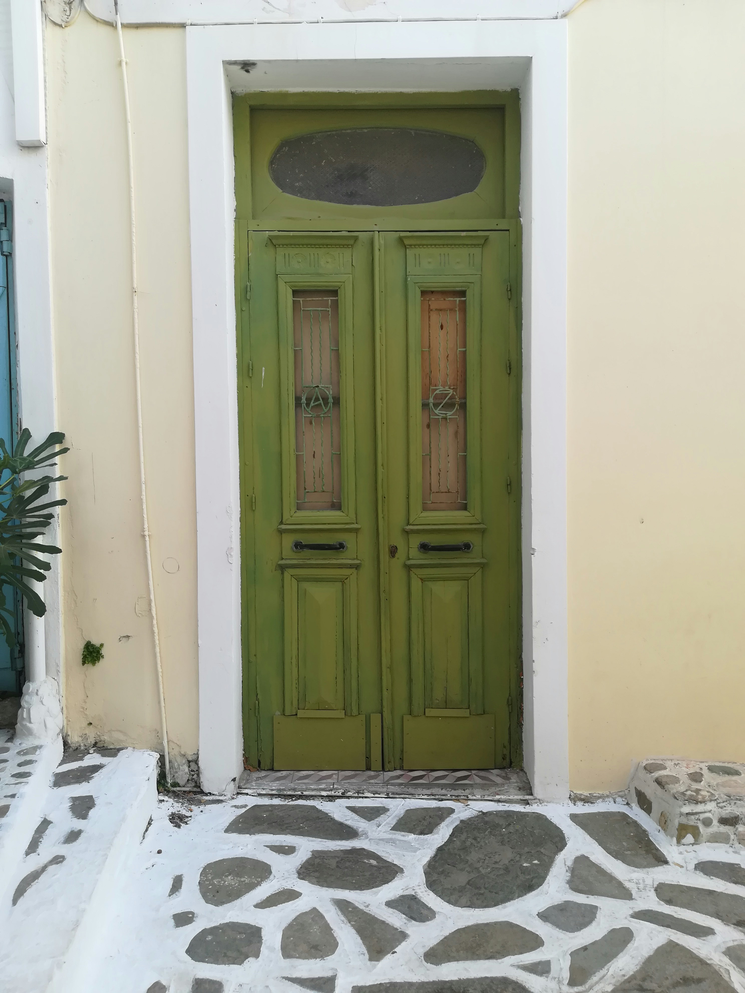 Olive-green double door set within a pale cream façade, framed by a white arch and a stone-paved foreground.