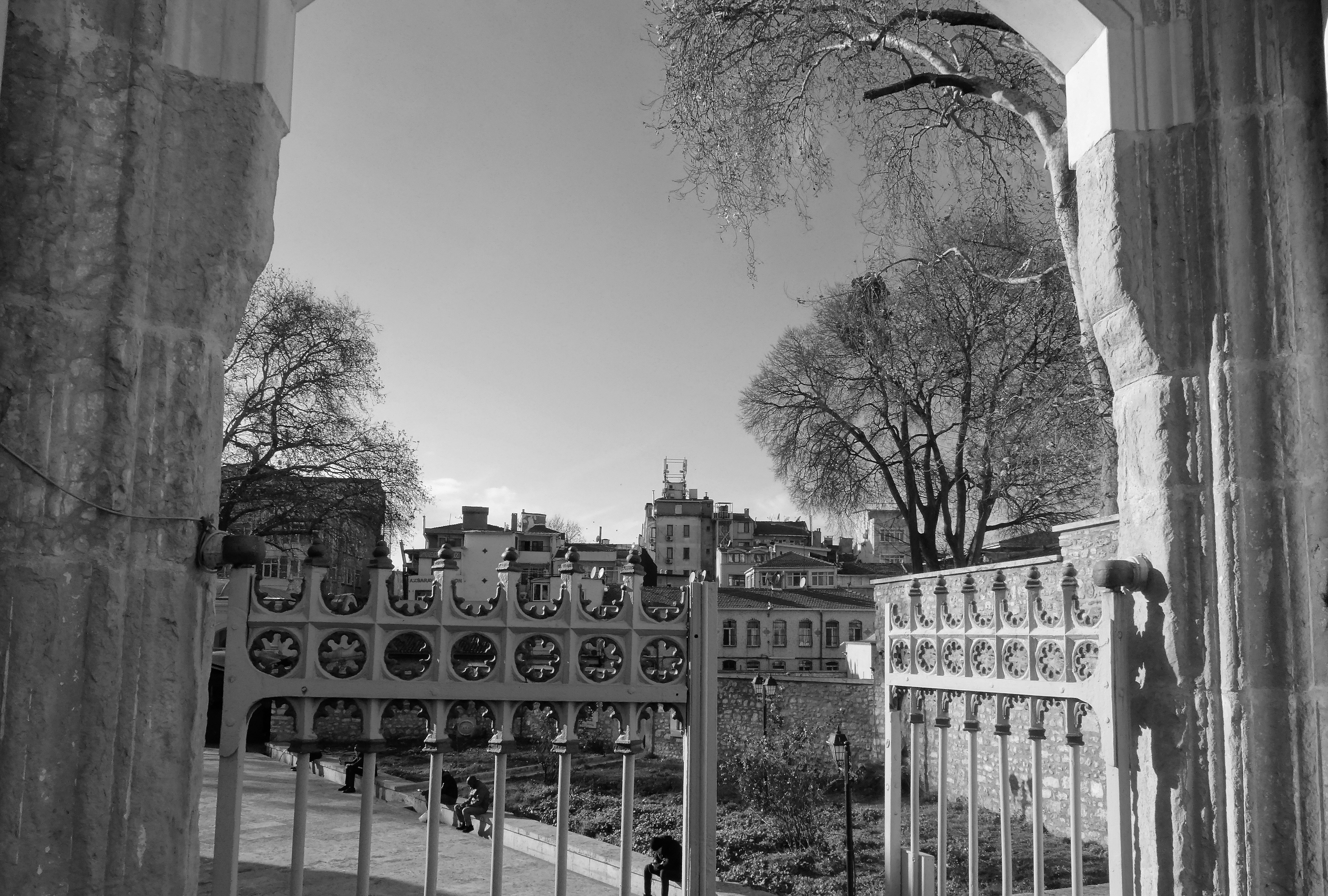 Stone archway framing an ornate gate and distant historic buildings under a clear sky.