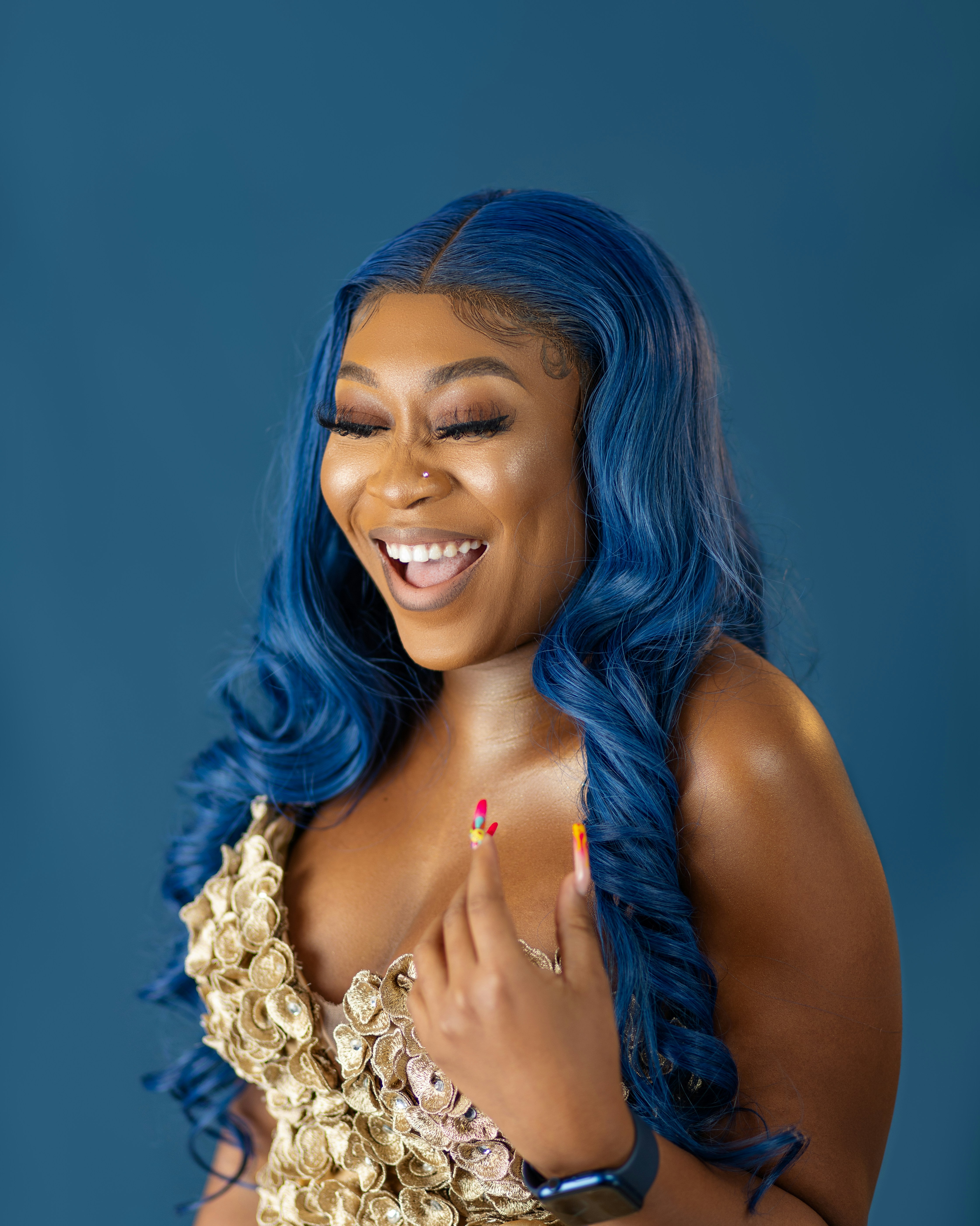 A woman with vibrant blue hair and a floral-patterned outfit expresses joy, set against a solid blue background.