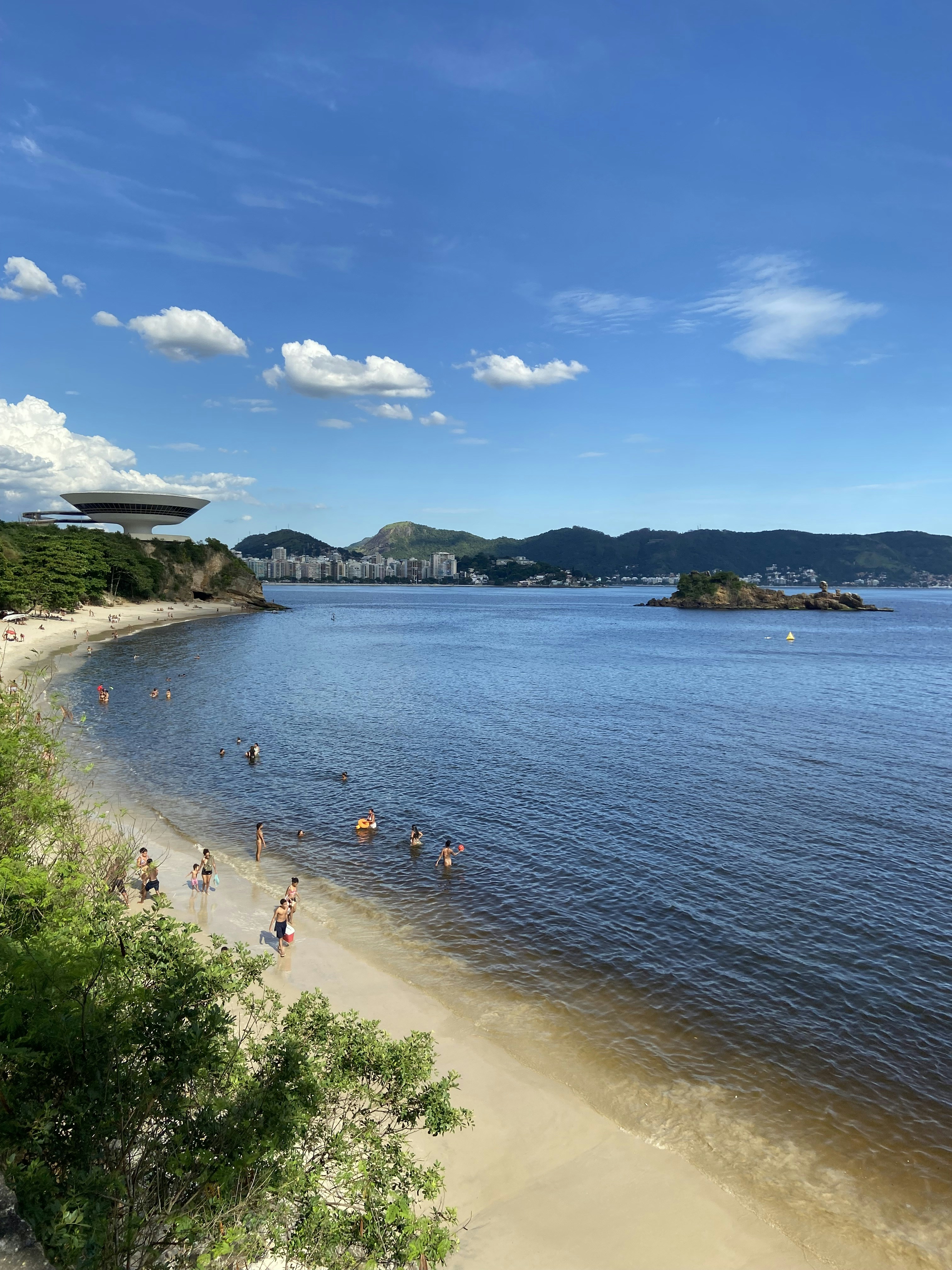 Beachgoers enjoying a sunny day along a tranquil shoreline, with cityscape and mountains in the background. A modern architectural structure overlooks the scene.