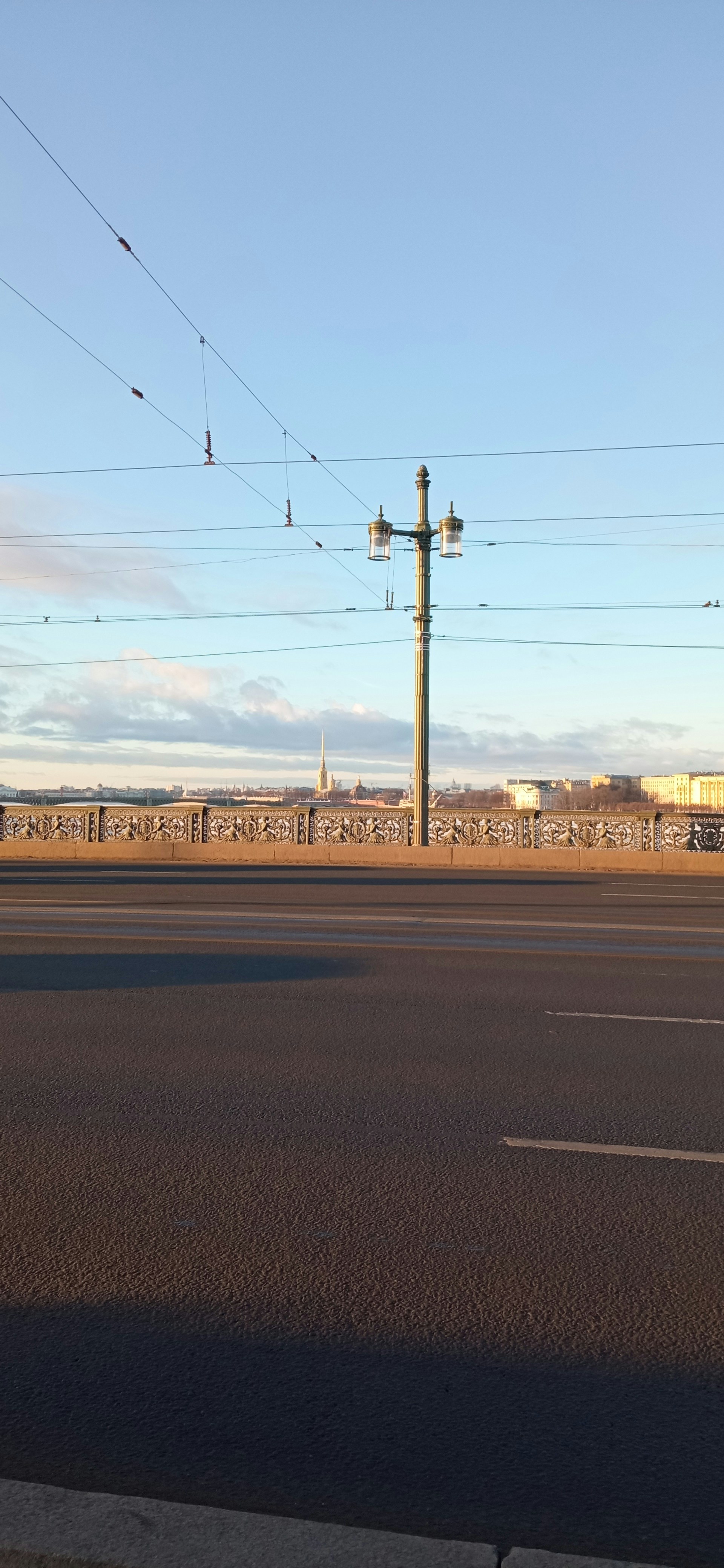 Street-level view along a bridge with decorative railing, a tall lamp post at center, and overhead tram wires crossing the scene. Distant city buildings line the horizon under a clear blue sky.