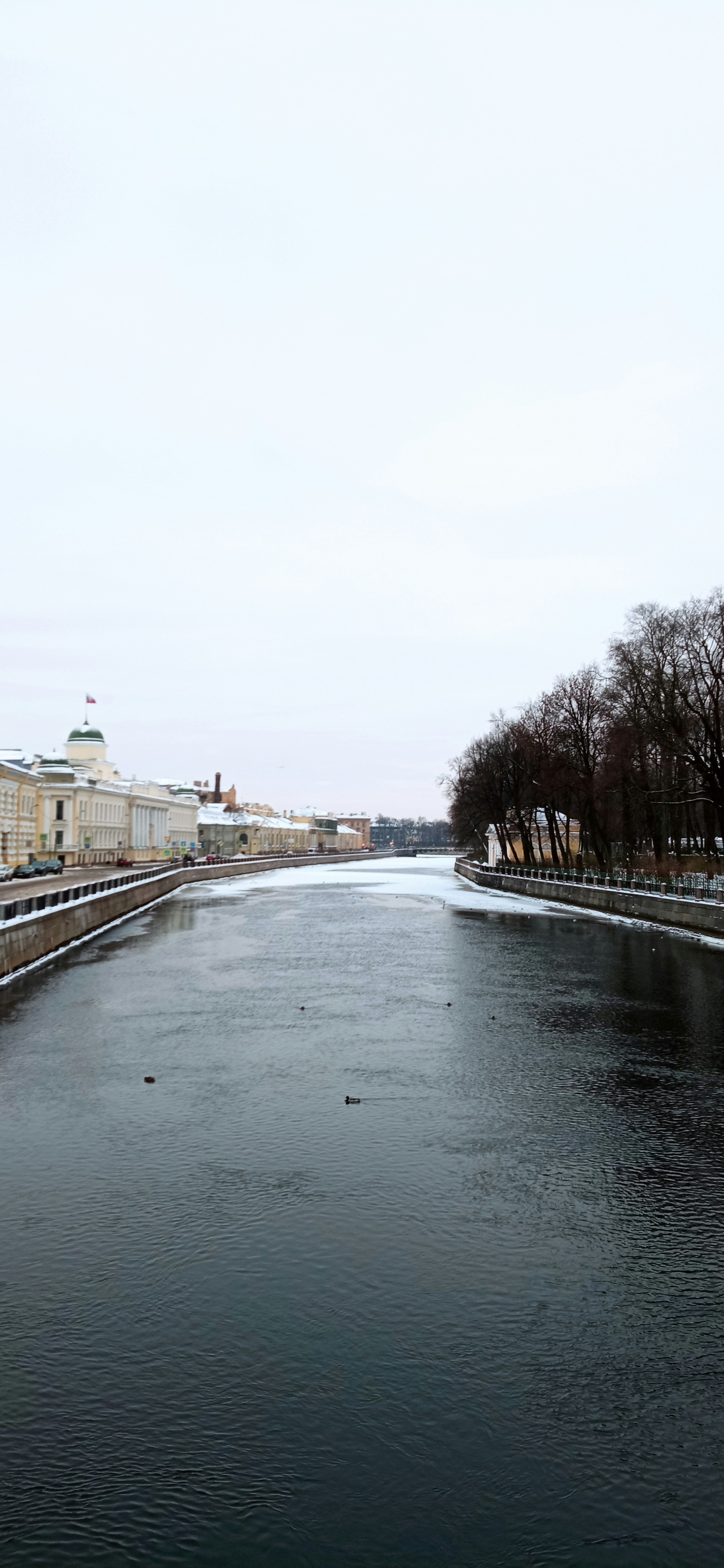Snow-tinted canal flanked by pale neoclassical buildings and bare trees, leading toward a distant city horizon. The calm water reflects the overcast sky, heightening the season's quiet mood.