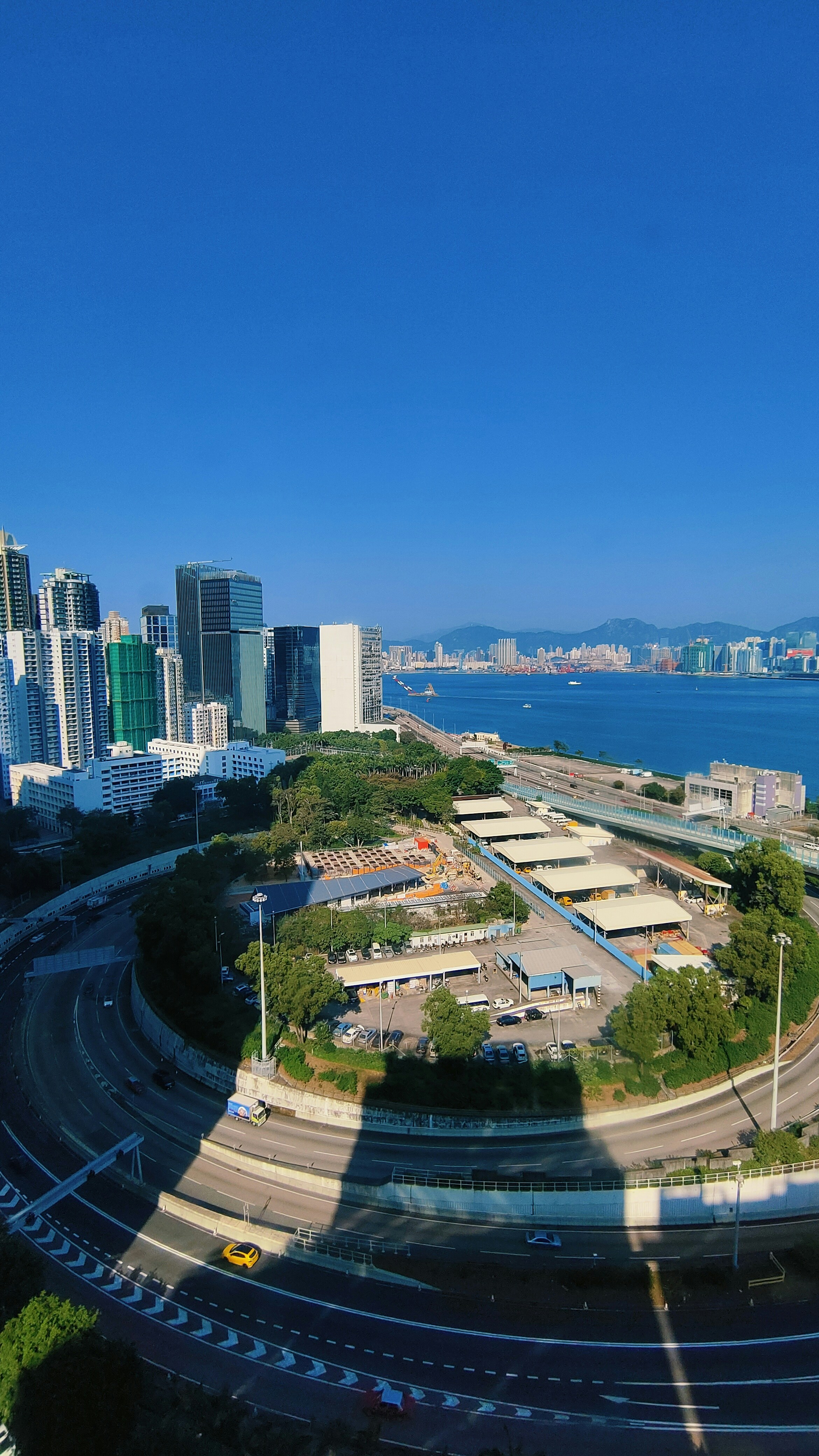 Aerial cityscape showing a cluster of high-rise buildings along a blue bay, with a curved ring road in the foreground casting long shadows. The photo emphasizes the urban silhouette against the water.