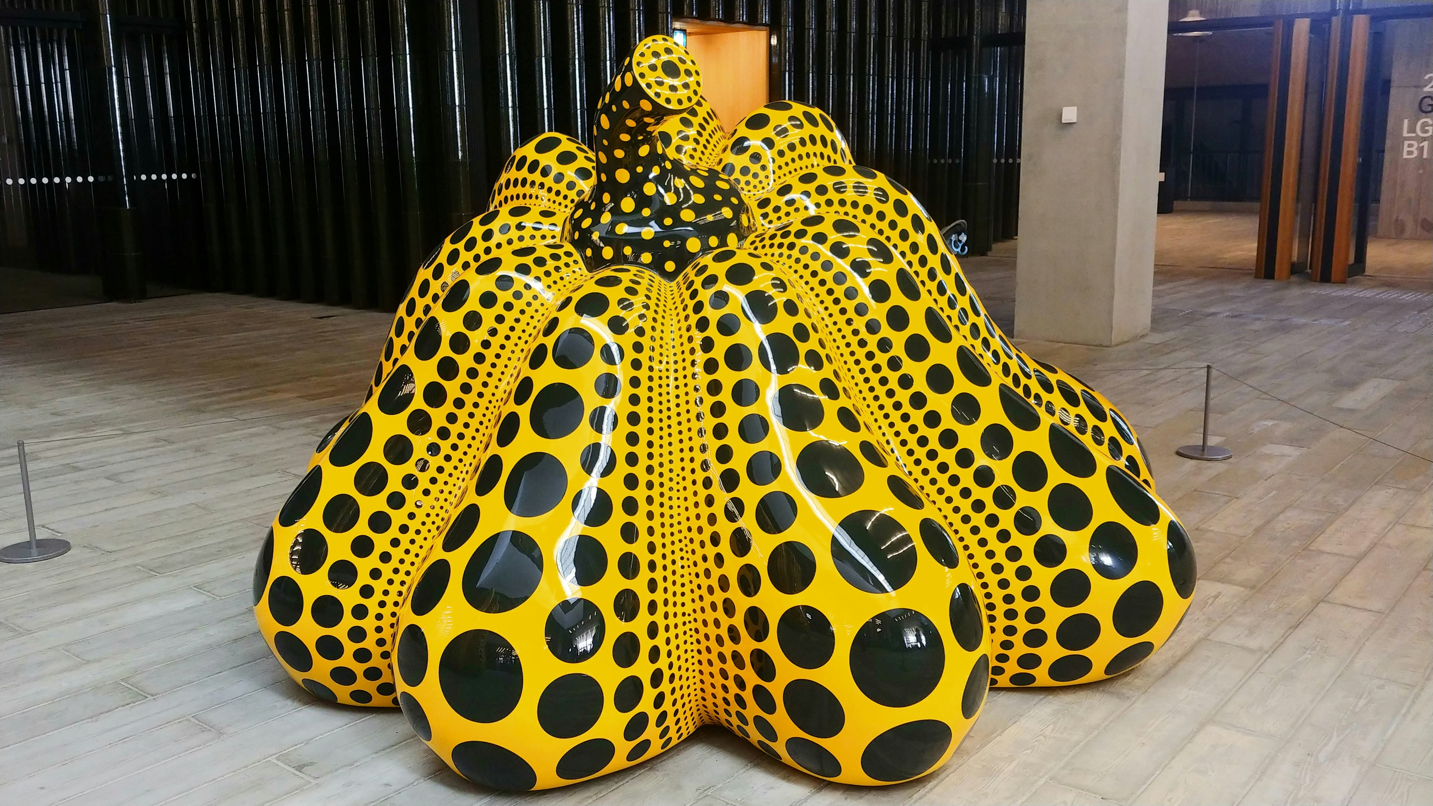 Kusama-inspired yellow pumpkin sculpture with black polka dots forms a clustered centerpiece in a modern gallery lobby.
