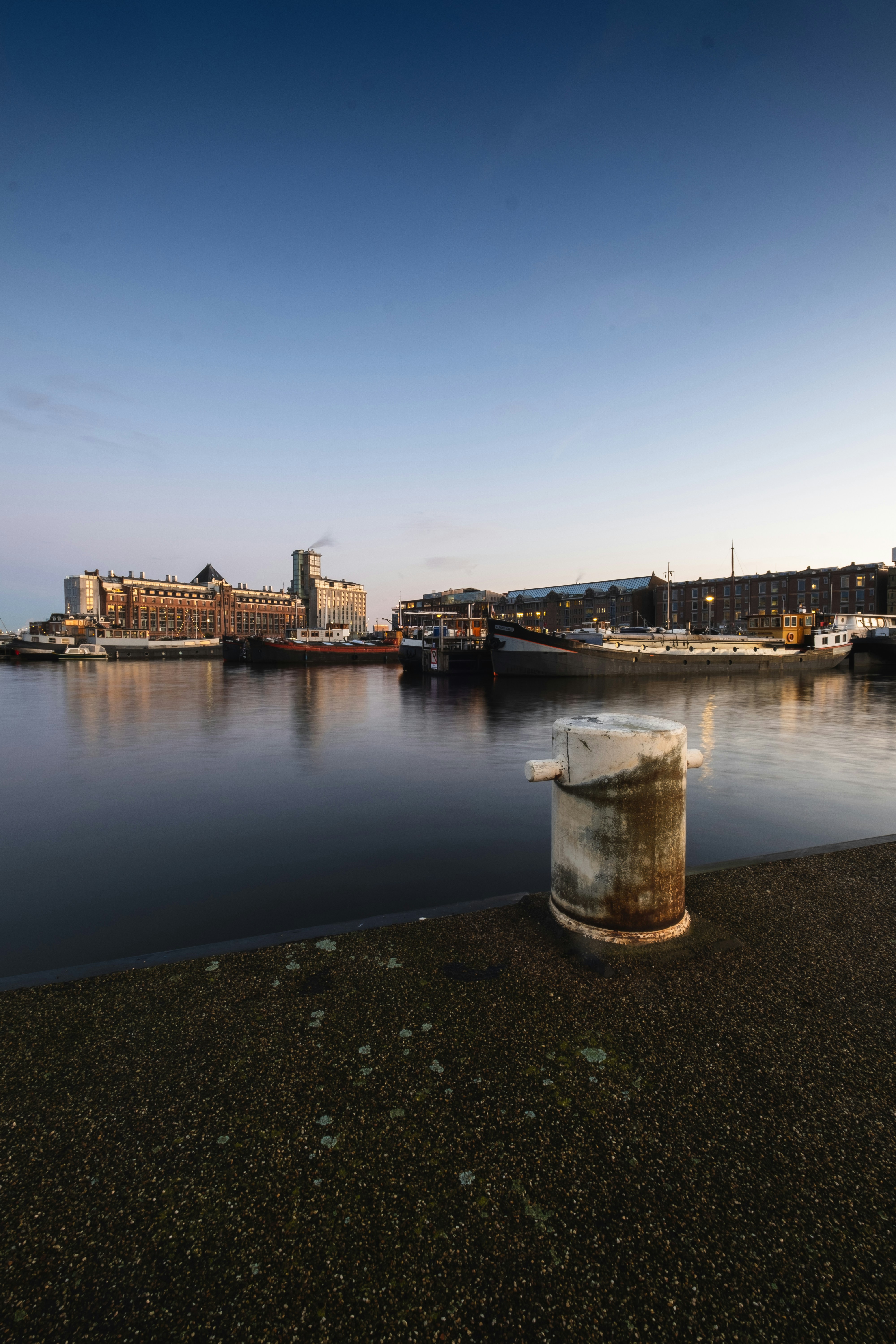This captivating image captures a tranquil waterfront scene at dusk, featuring a dockside bollard in the foreground and a line of moored boats and charming buildings in the background. The composition skillfully balances the textured surface of the dock with the smooth, reflective waters, while the sky transitions from a warm golden hue to deep blue, enhancing the serene atmosphere. The soft lighting and subtle color palette create a peaceful mood, making the image visually striking and evocative.