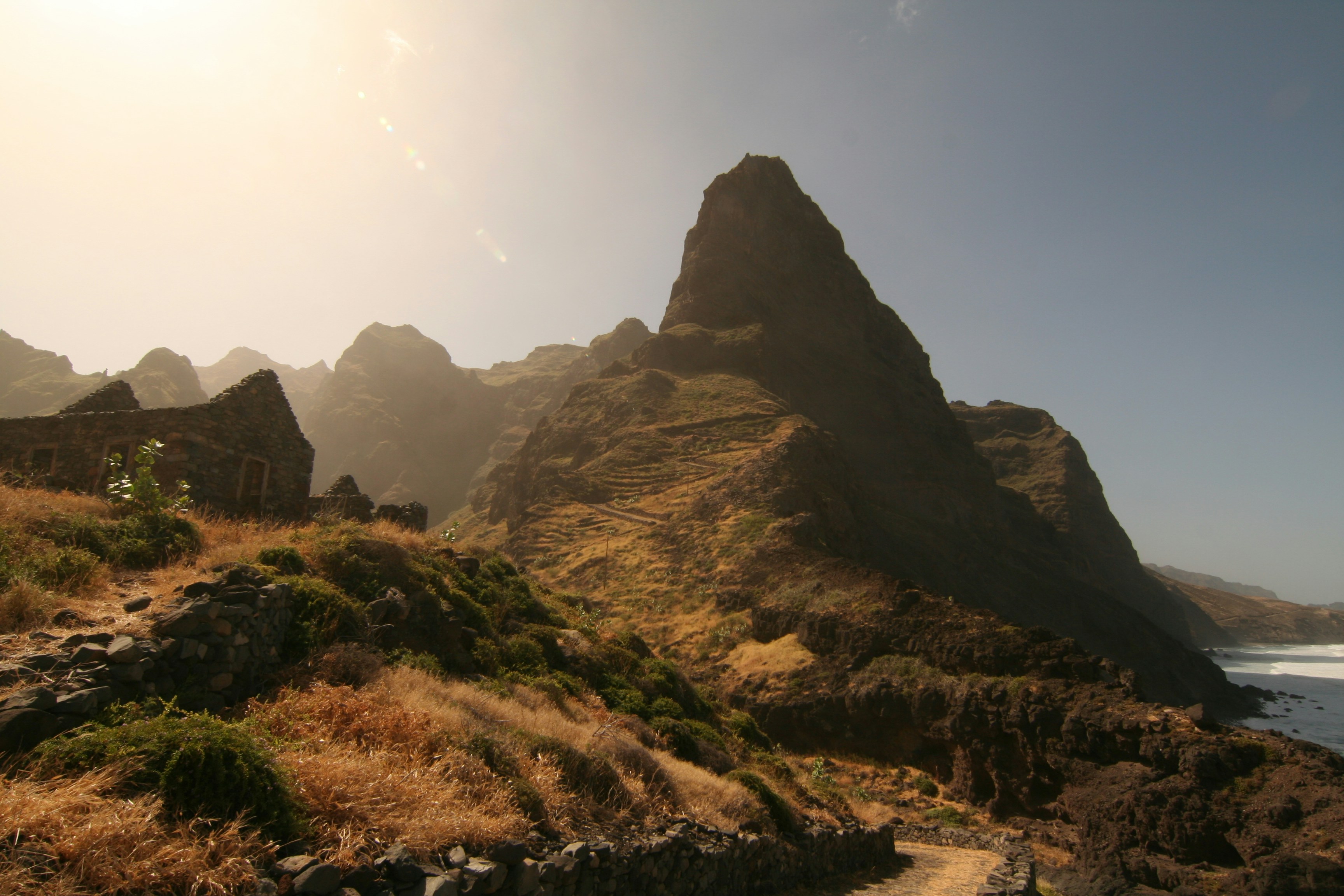 Rugged mountain landscape under a warm, hazy sky with a path winding through dry grass and rocky terrain.