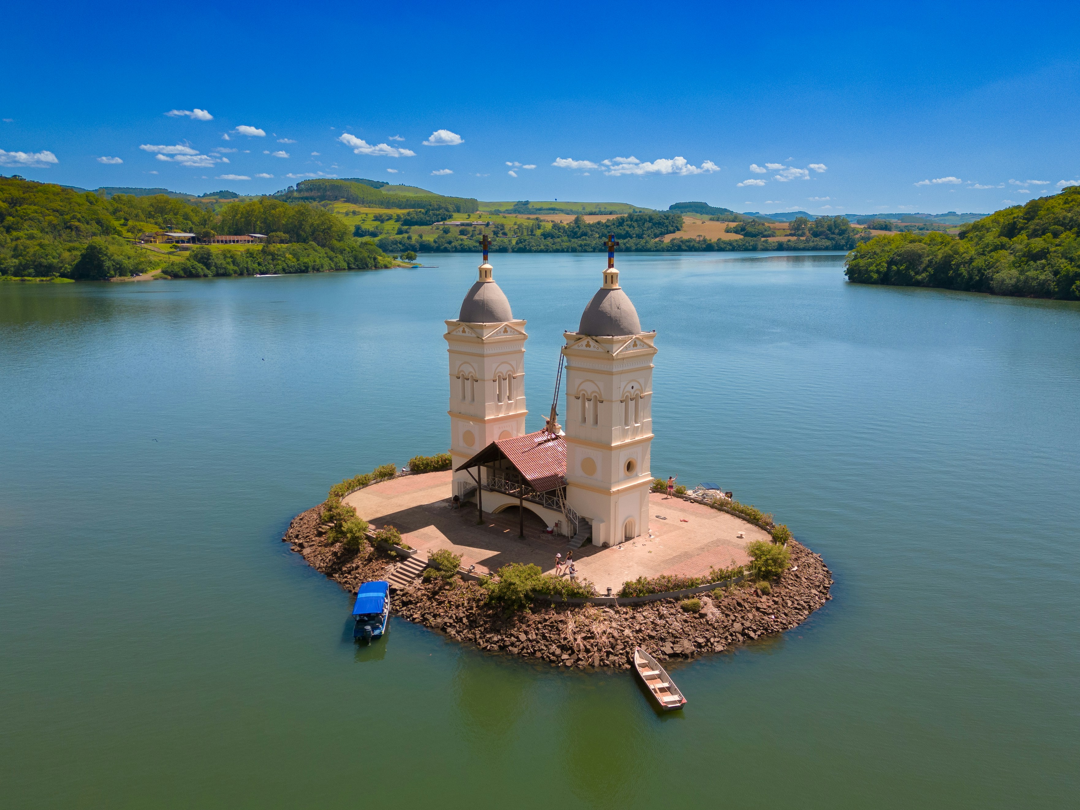 Twin stone towers rise from a small island surrounded by a vast, calm lake under a clear blue sky.
