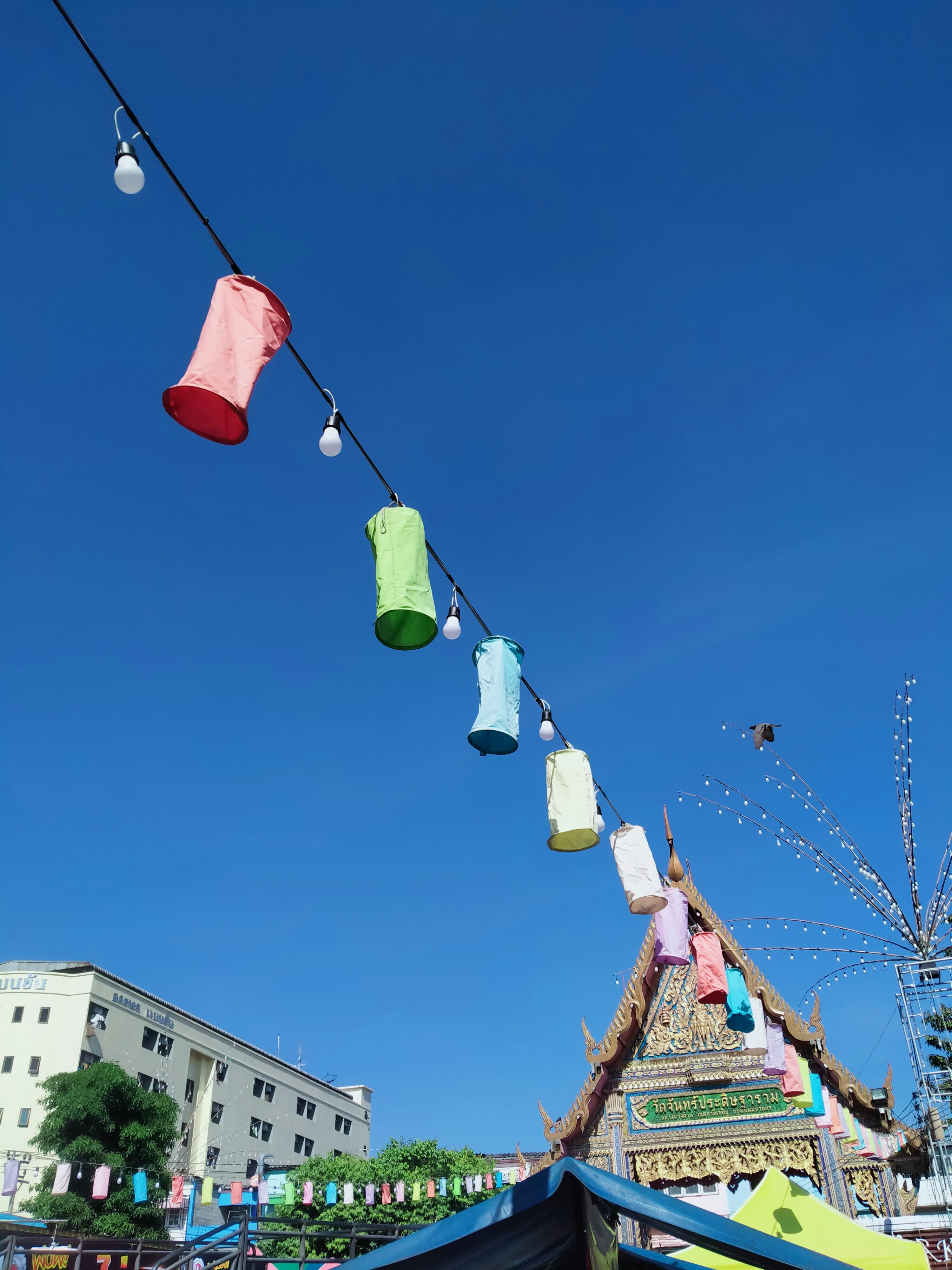 Colorful lanterns hang in a diagonal line across a clear blue sky above a temple festival square. Tents and festival structures populate the foreground, highlighting a lively outdoor scene.