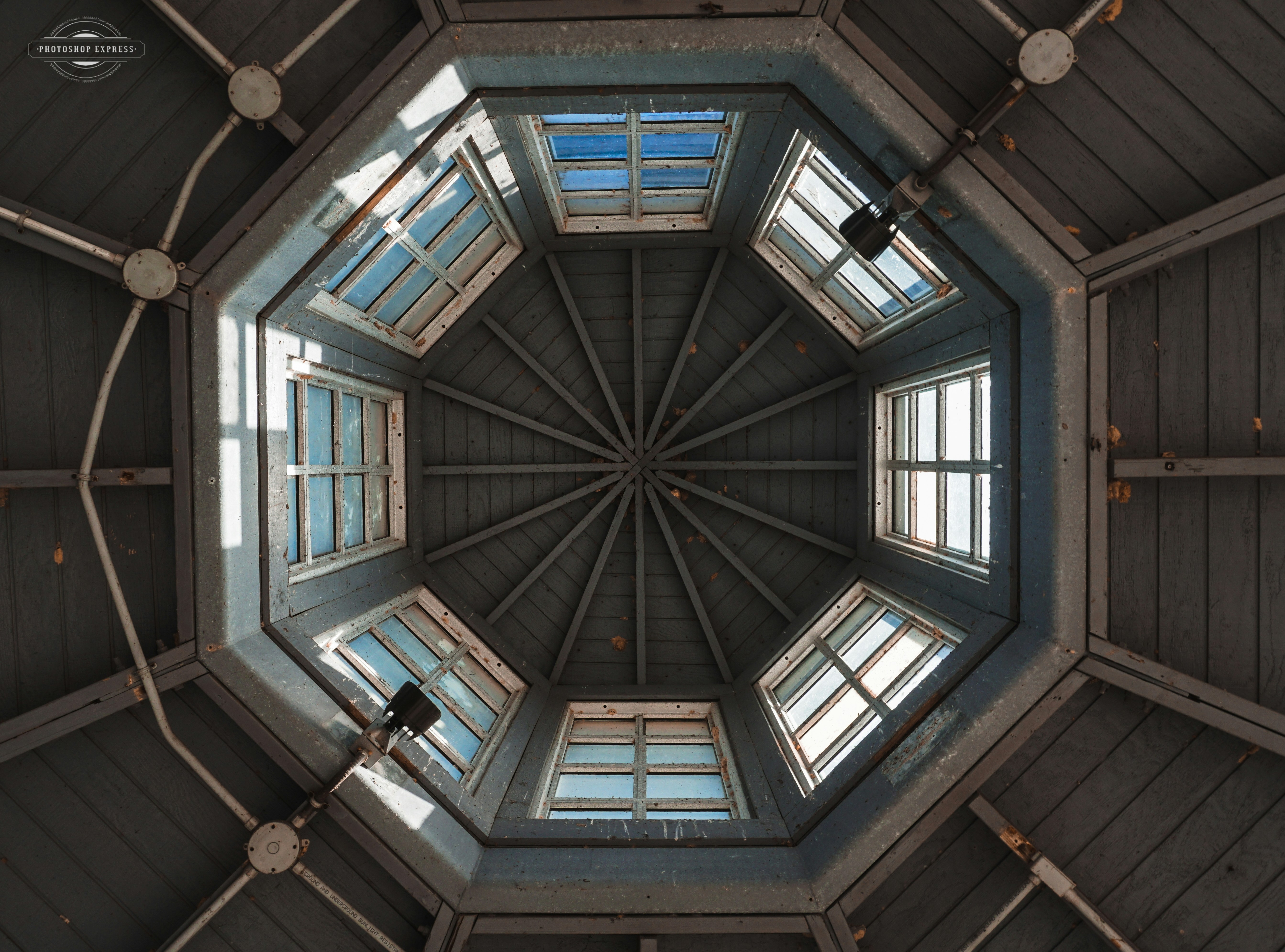 View of an octagonal ceiling with multiple windows, showcasing intricate wooden beams and a clear blue sky above.