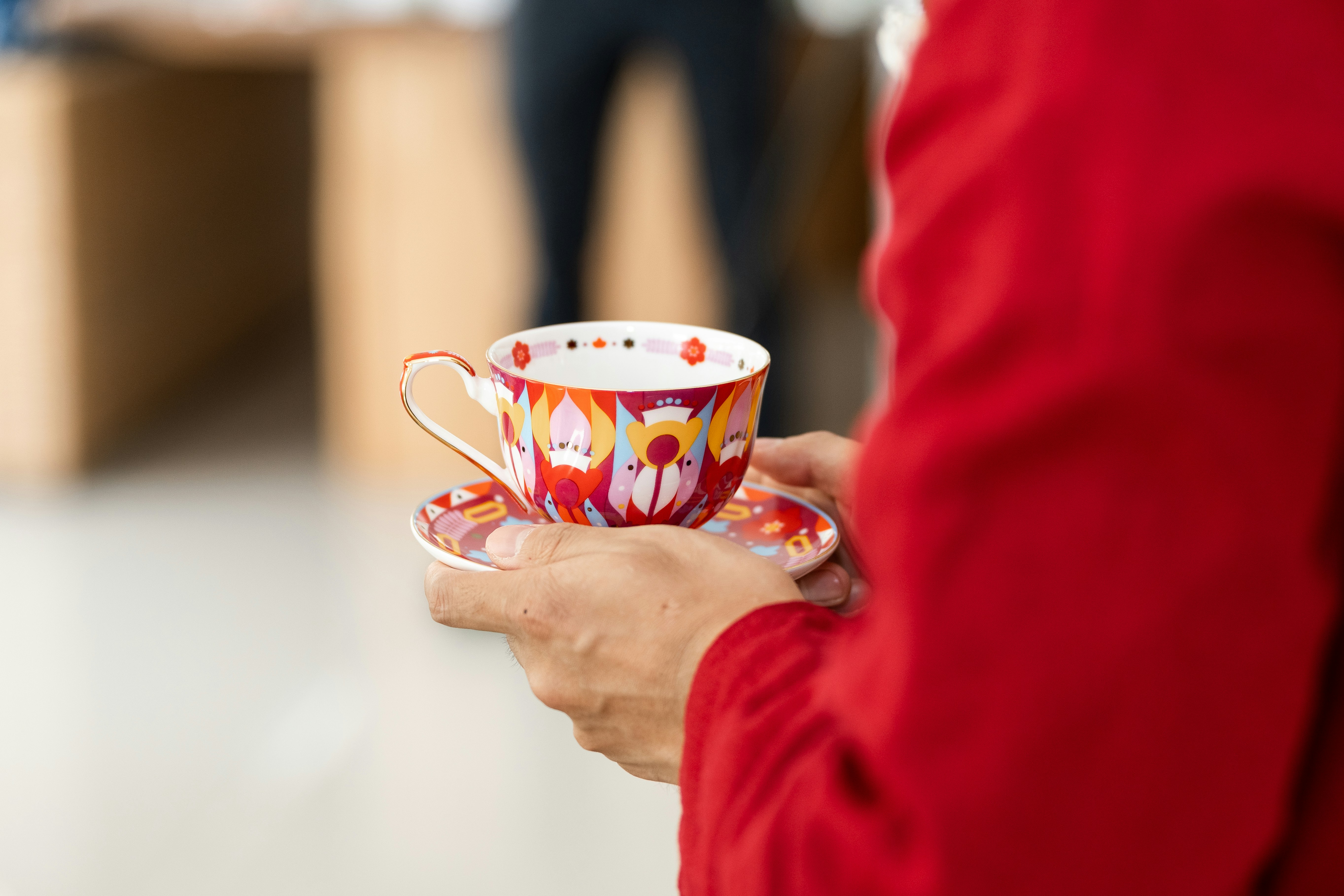 Colorful floral teacup and saucer held delicately by a person in a red shirt, showcasing a blend of artistry and warmth.