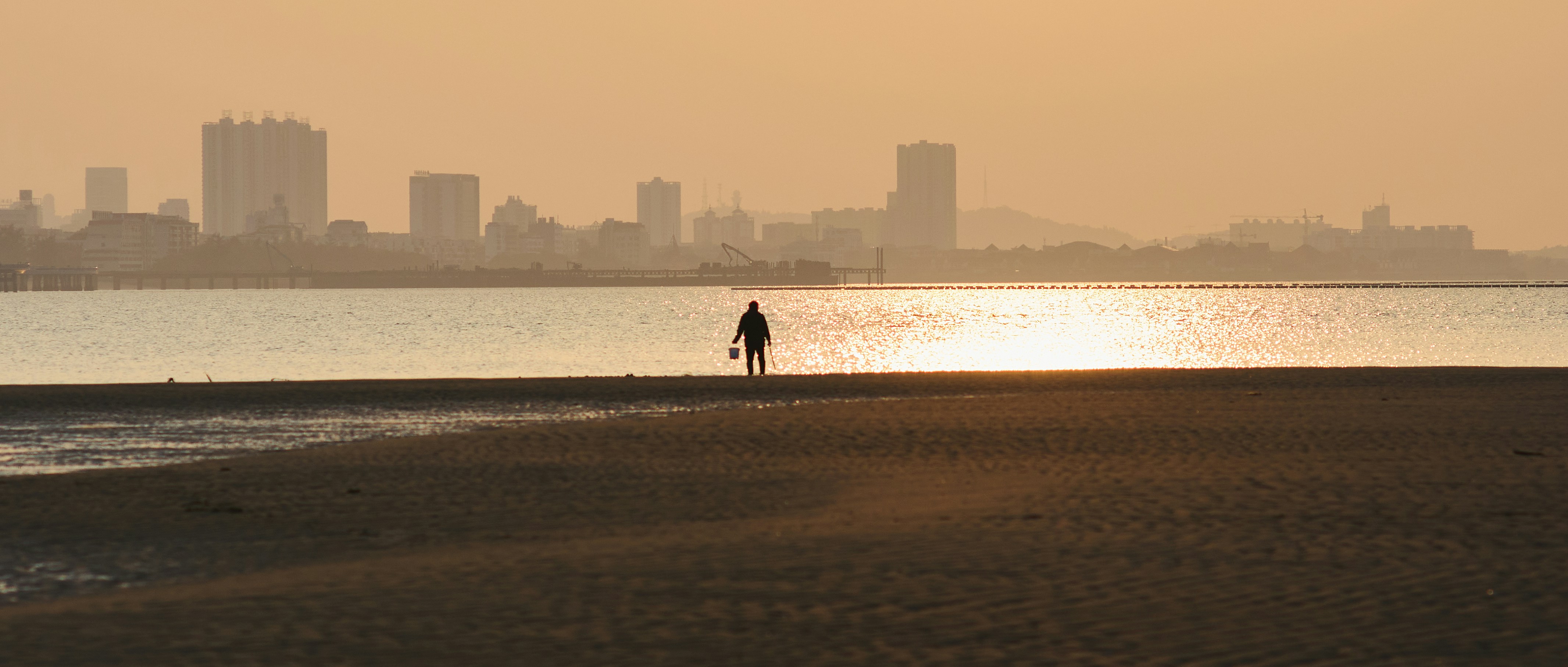 Silhouette of a person walking along a beach with city skyline in the background at sunset.