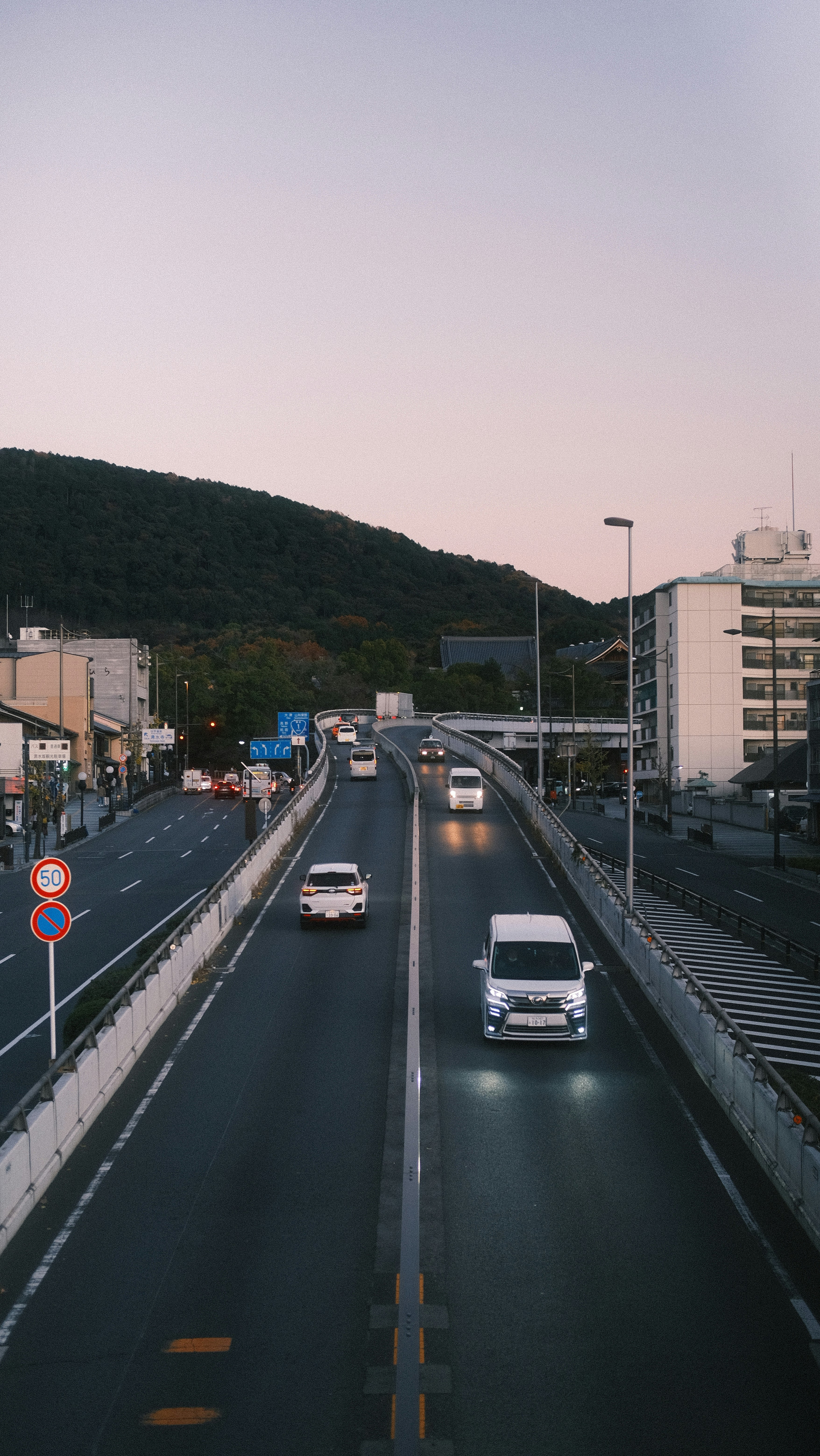 Cars navigating a winding road under a twilight sky, surrounded by urban buildings and a distant hillside.