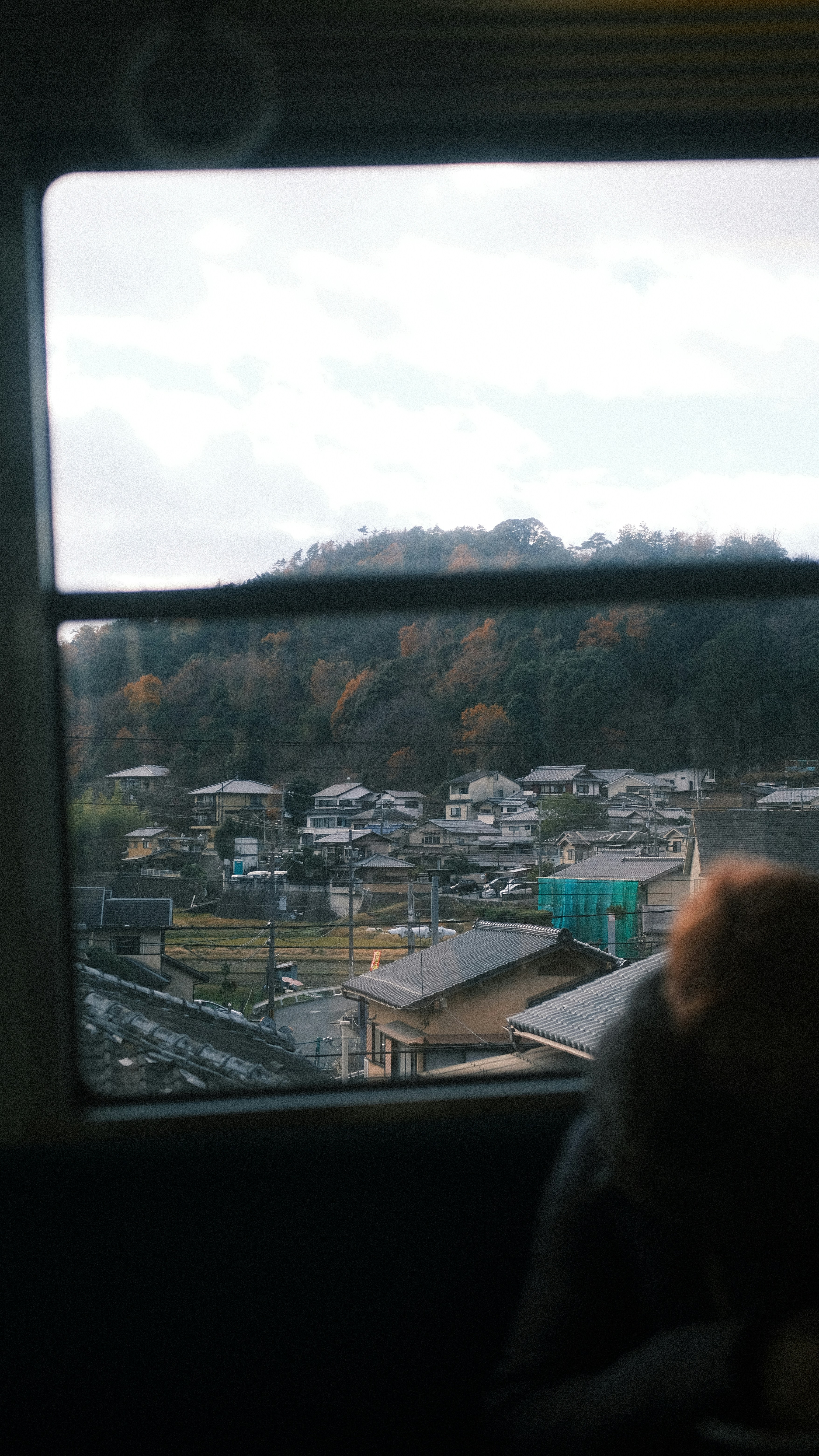 View of a serene town nestled among hills, framed by a train window. The scene captures the essence of rural life with autumn foliage in the background.