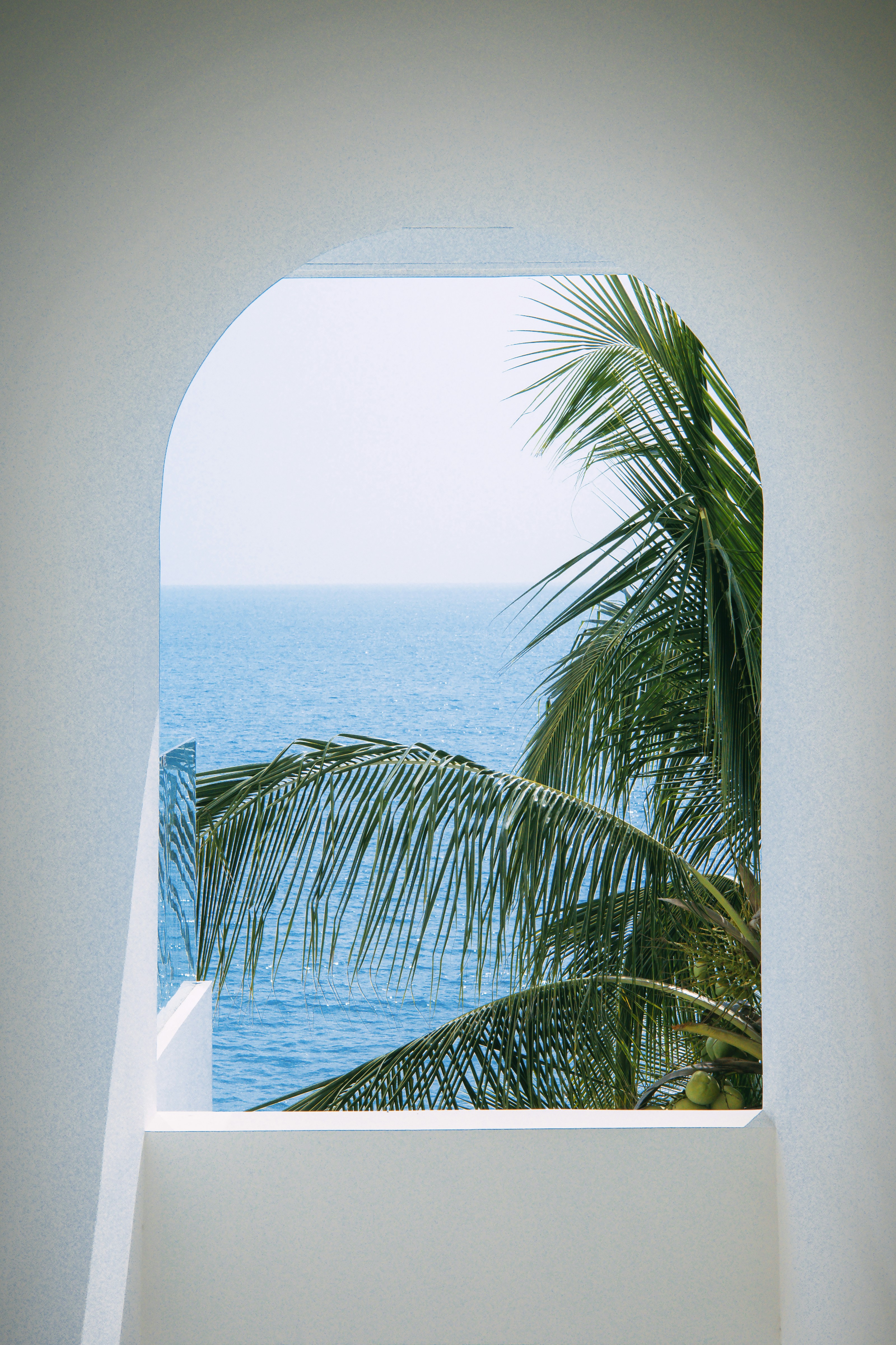 View through an arched opening showcasing a tranquil sea and swaying palm fronds. The scene evokes a sense of peace and relaxation.