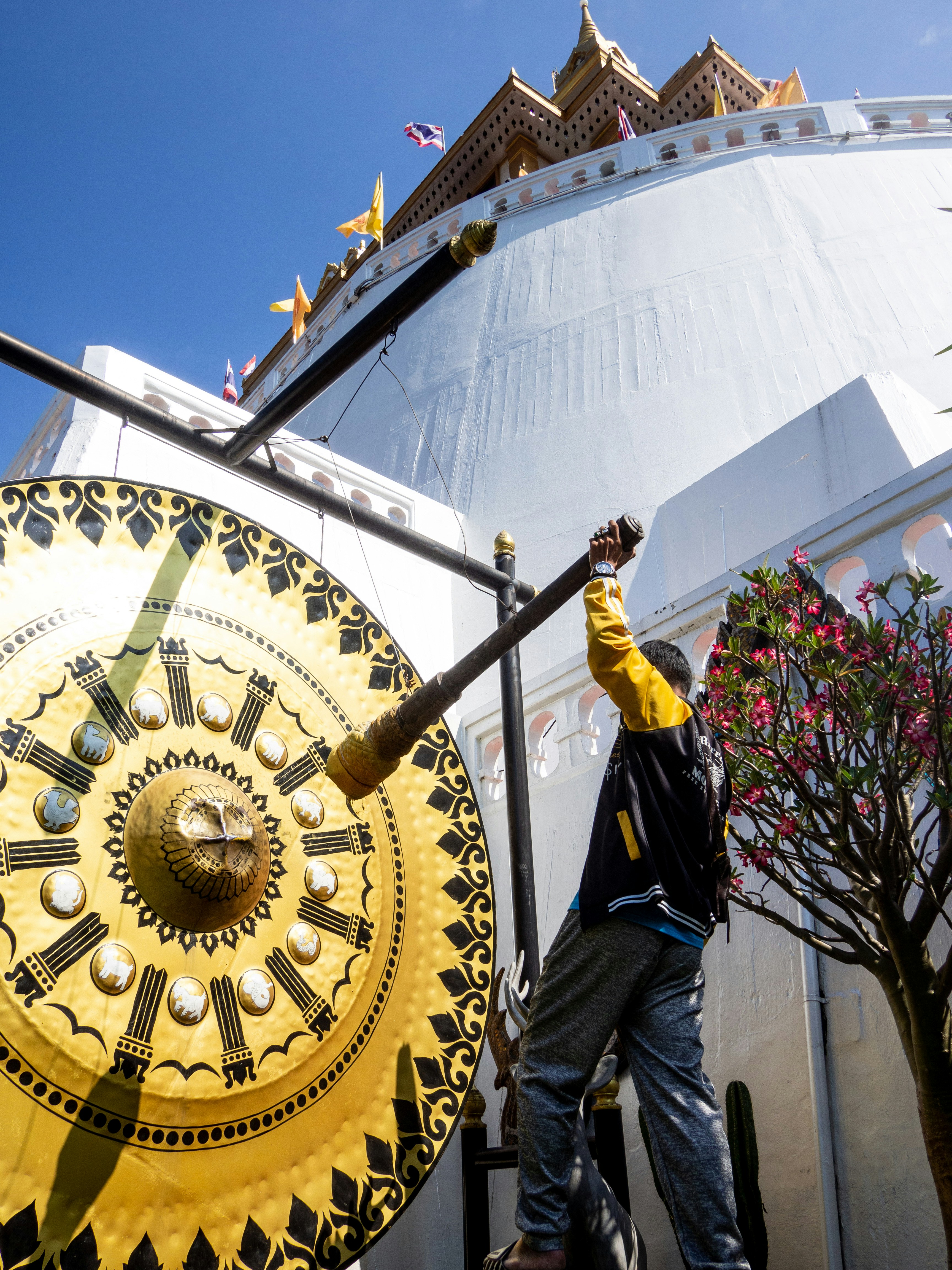 Person striking a large decorative gong beneath a grand stupa with flags fluttering against a clear blue sky.