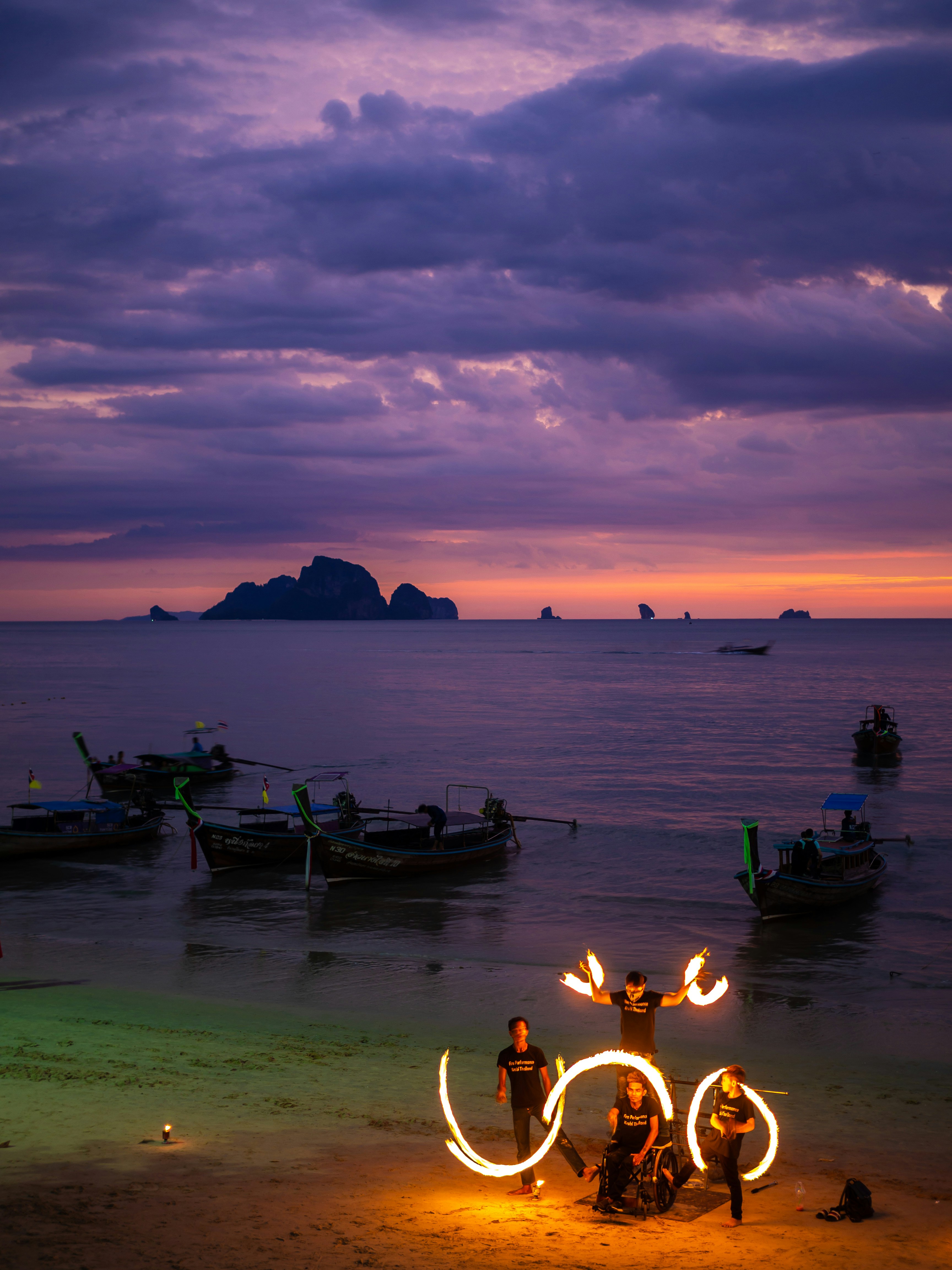 Fire performers create mesmerizing trails of light on the beach as twilight descends, with silhouettes of fishing boats in the background.