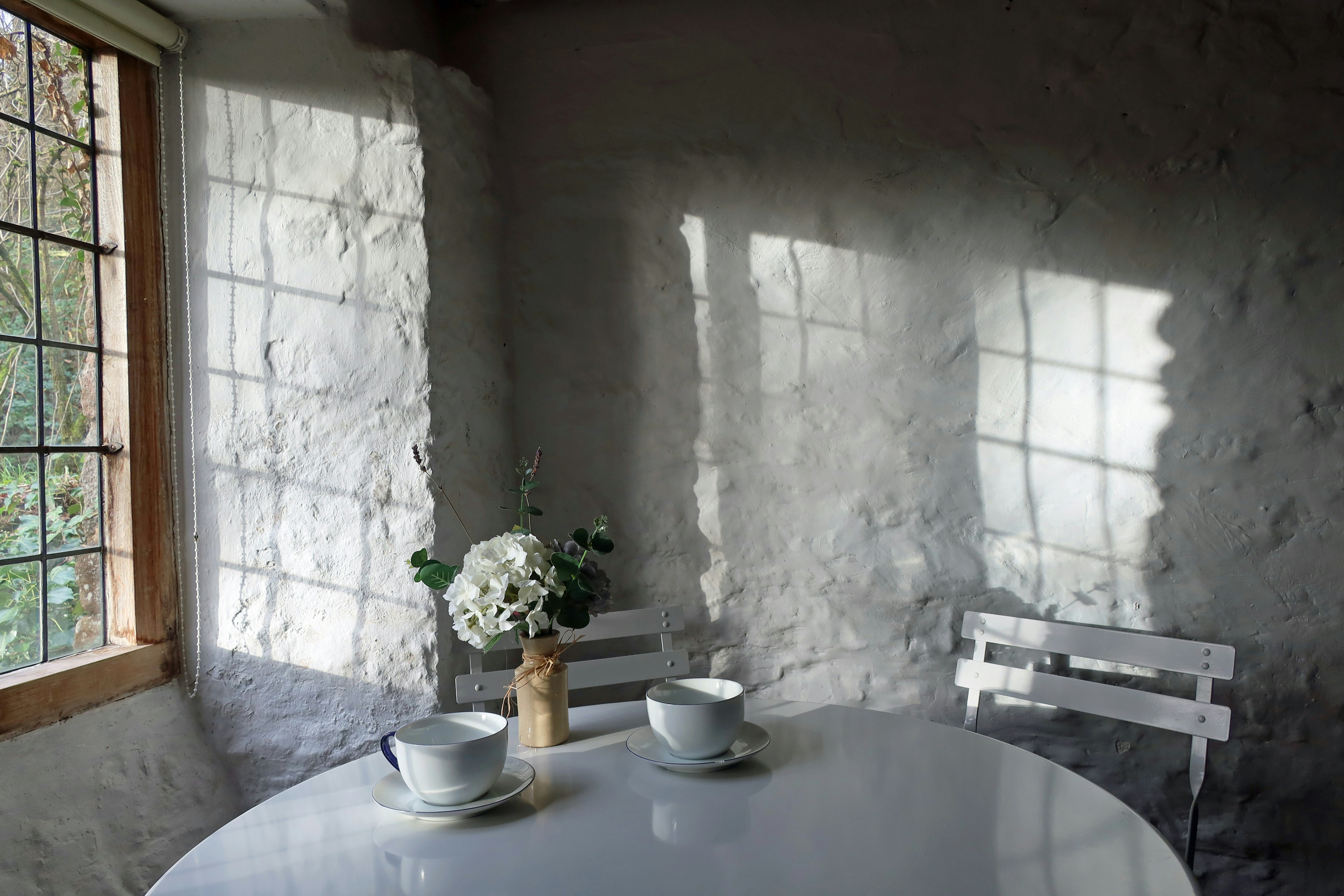 A cozy table setting with two cups and a vase of flowers, illuminated by soft shadows cast through a window onto textured walls.