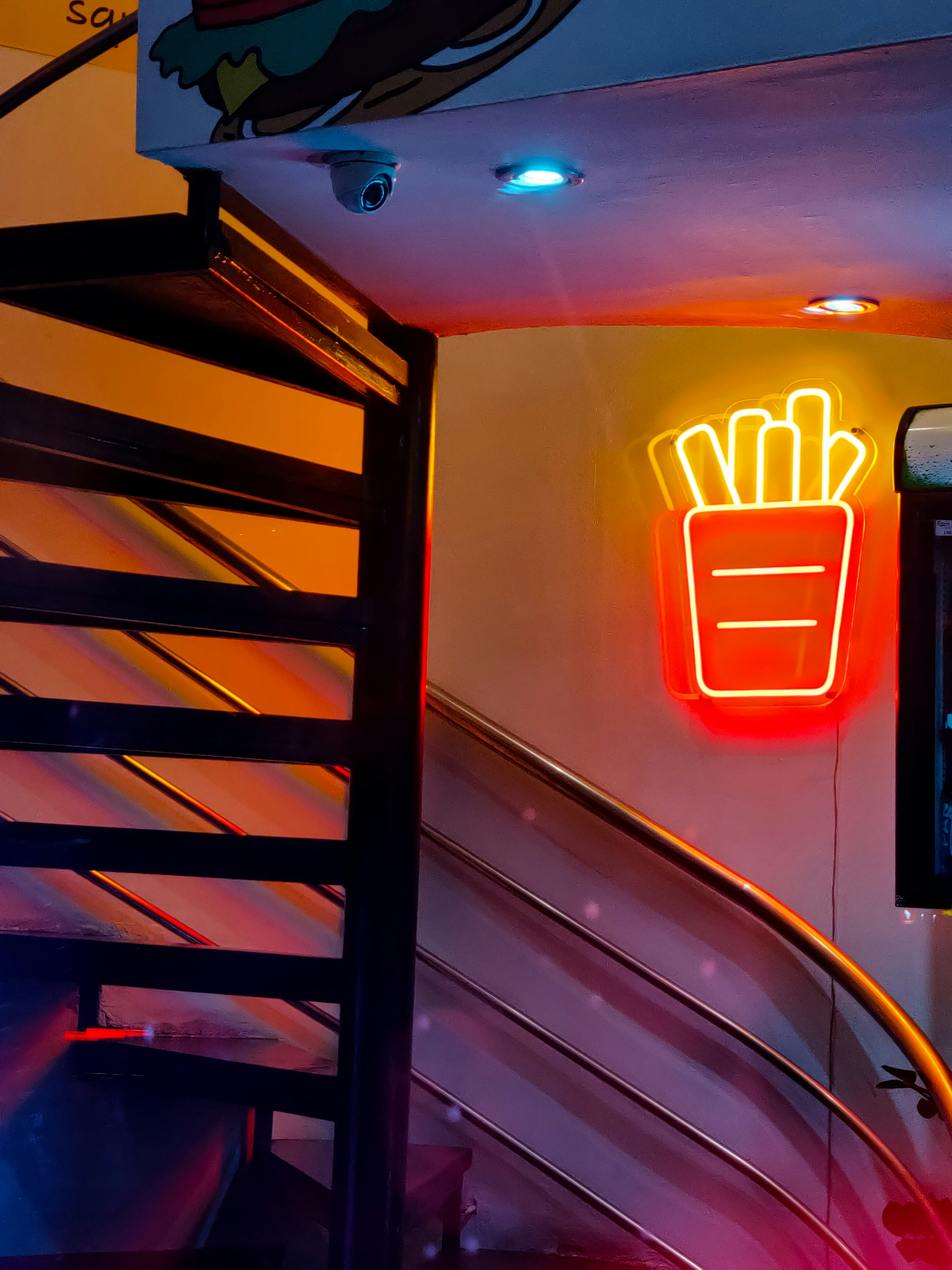 Neon fries sign glowing on a wall next to a spiral staircase, creating a vibrant atmosphere in a casual dining space.