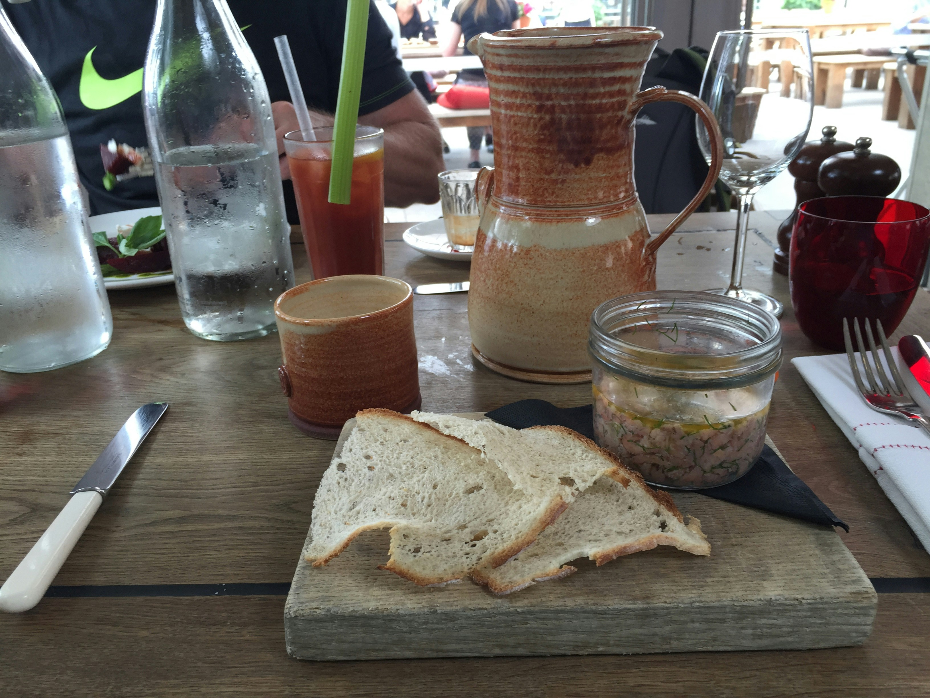 Sliced bread rests on a weathered wooden board at the center of a café table, with a ceramic pitcher, glassware, and jars nearby.