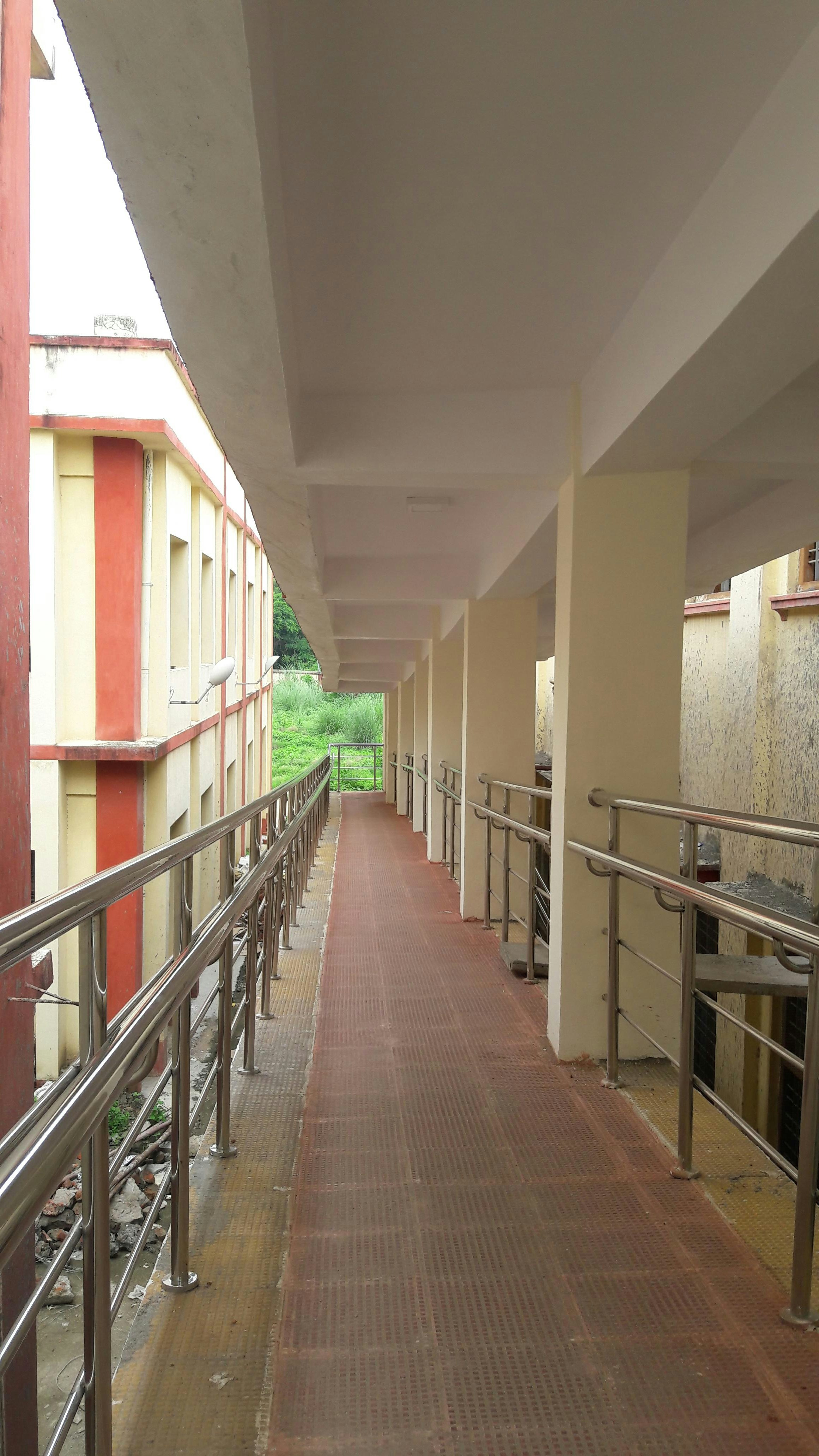 Long corridor flanked by buildings, featuring a metal railing and textured flooring, leading towards a green landscape in the background.