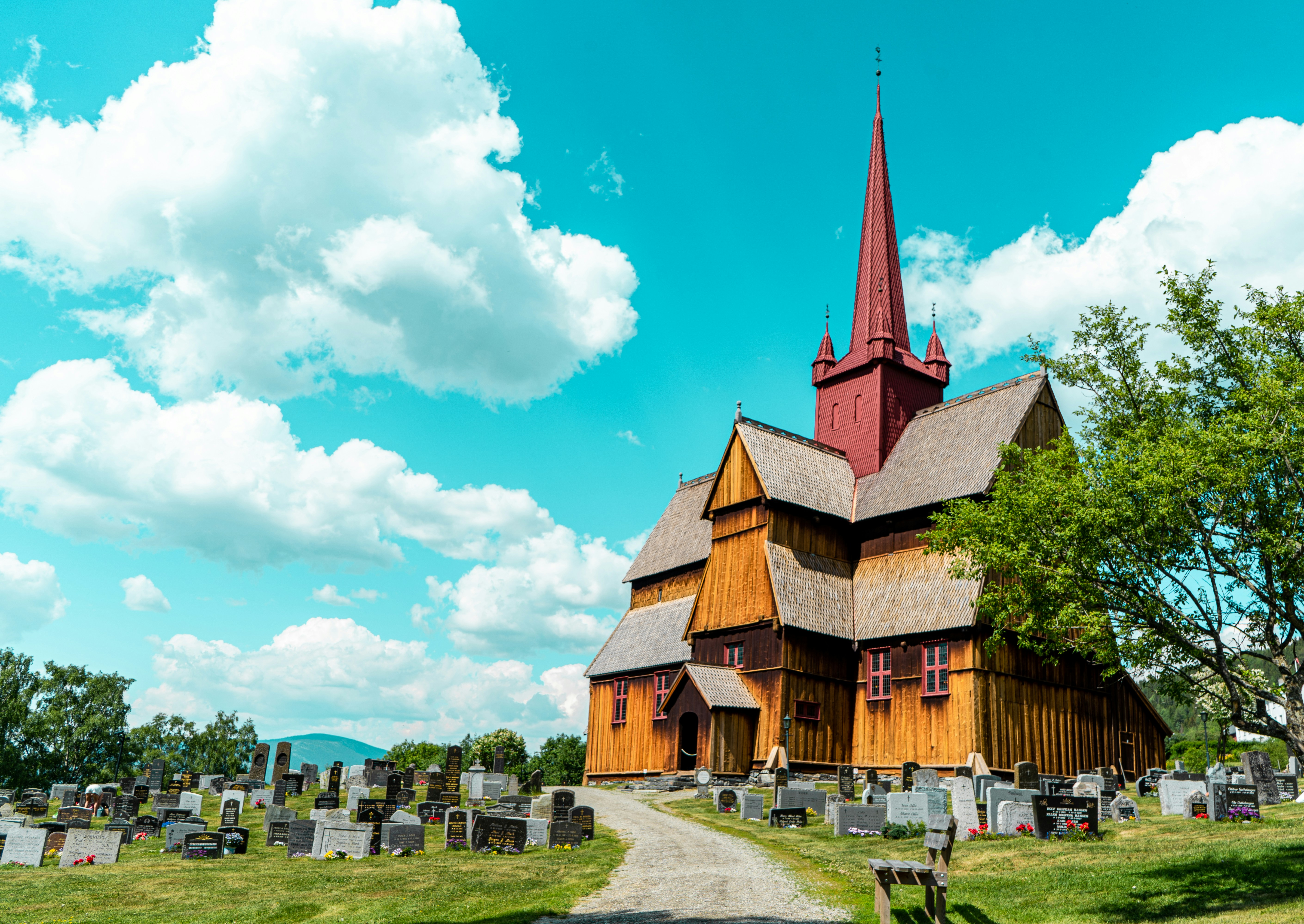 A striking wooden church with a tall red spire stands prominently in a cemetery, surrounded by gravestones under a bright blue sky dotted with clouds.
