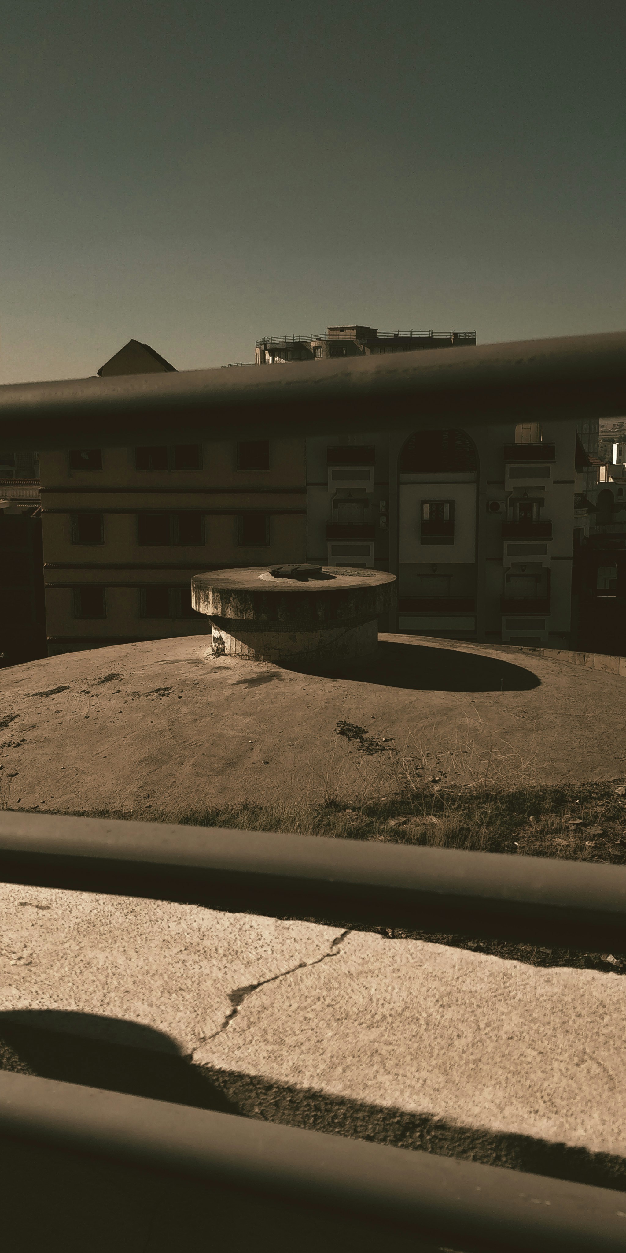 An abandoned circular platform on a rooftop, surrounded by urban buildings under a clear sky. The scene evokes a sense of neglect and solitude.