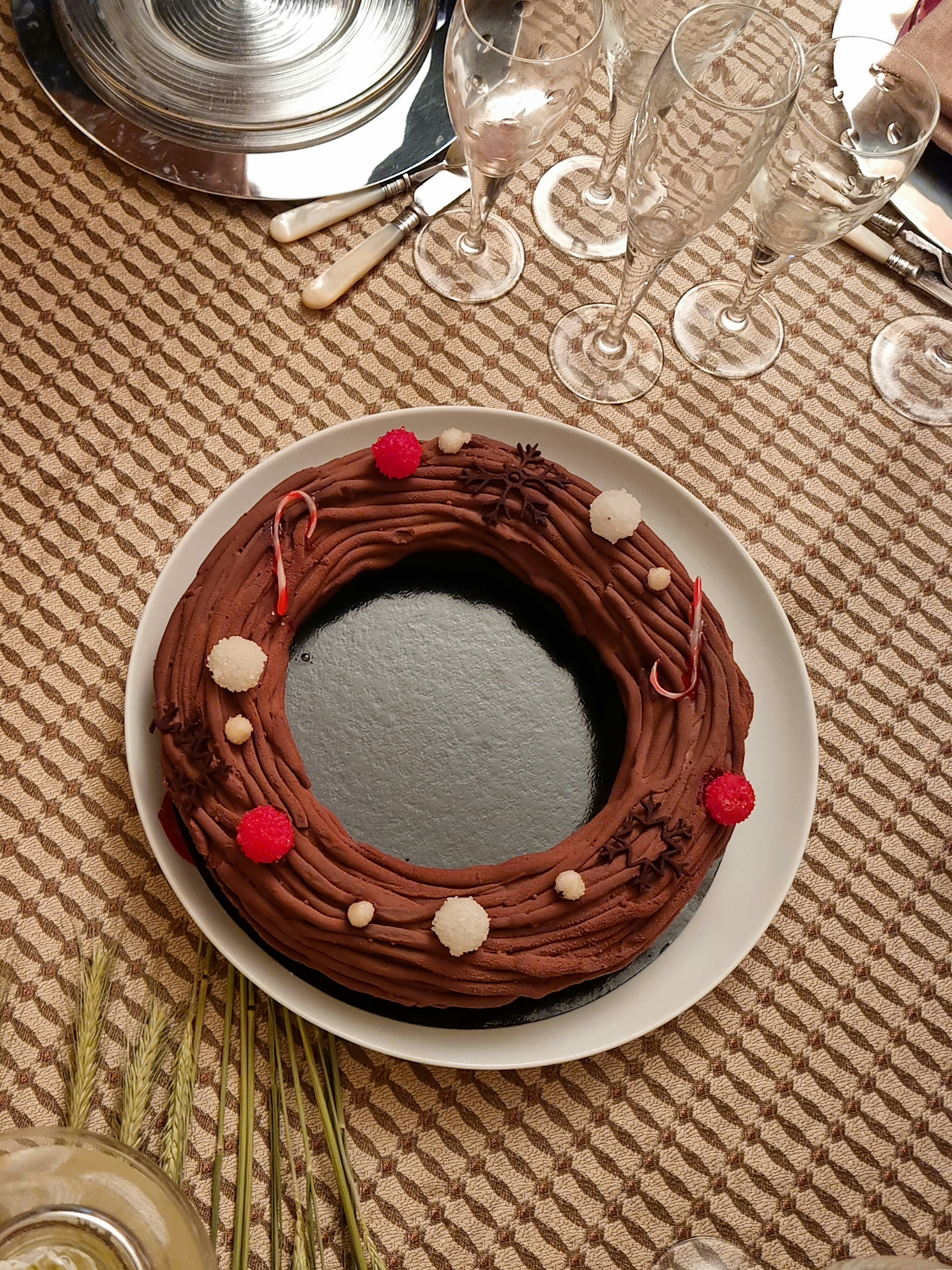 Chocolate wreath cake adorned with festive decorations, surrounded by elegant glassware on a textured tablecloth.