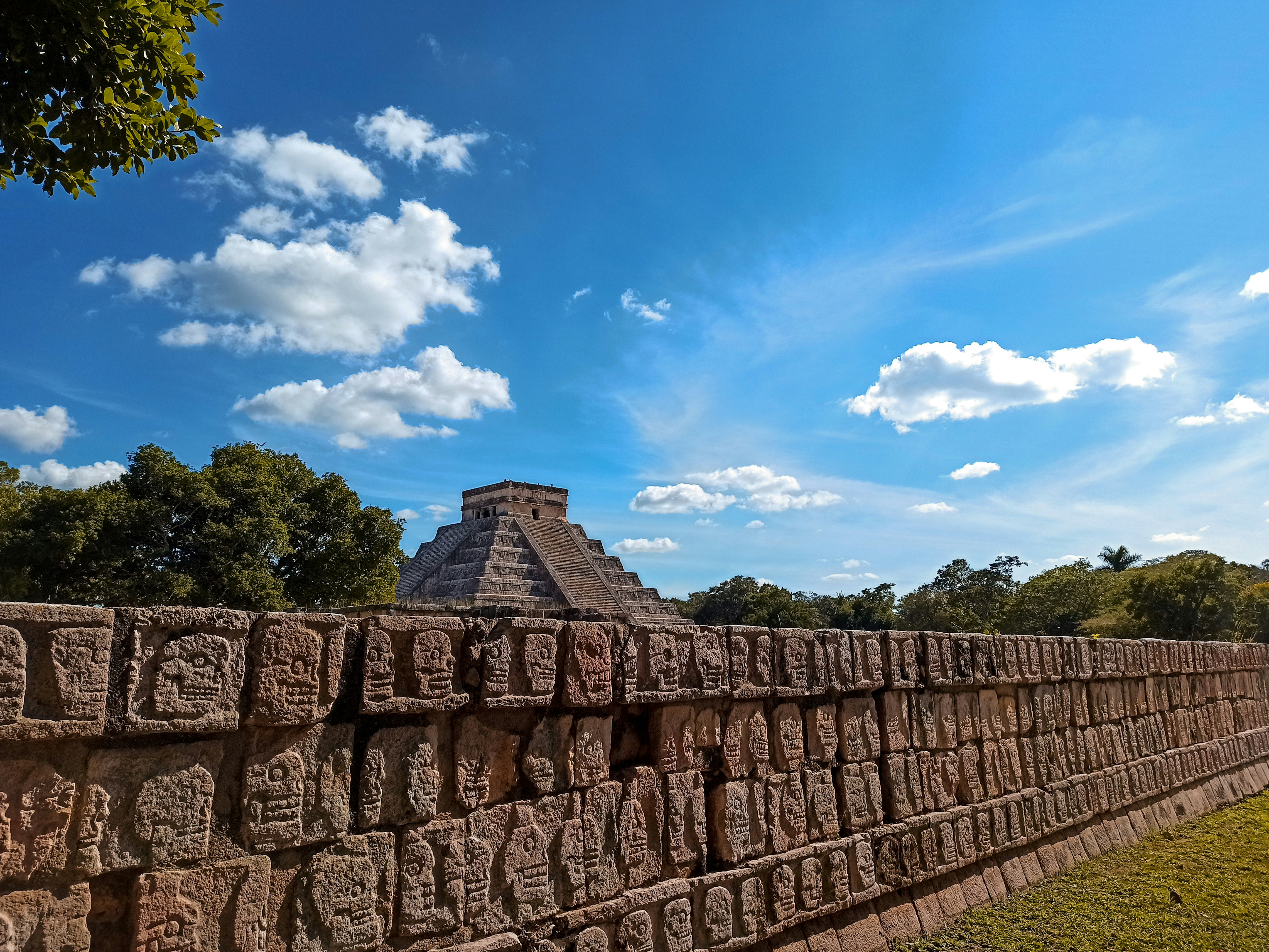 A historic pyramid rises majestically behind a carved stone wall, framed by a vibrant sky dotted with clouds. The scene captures the essence of ancient architecture in a lush landscape.