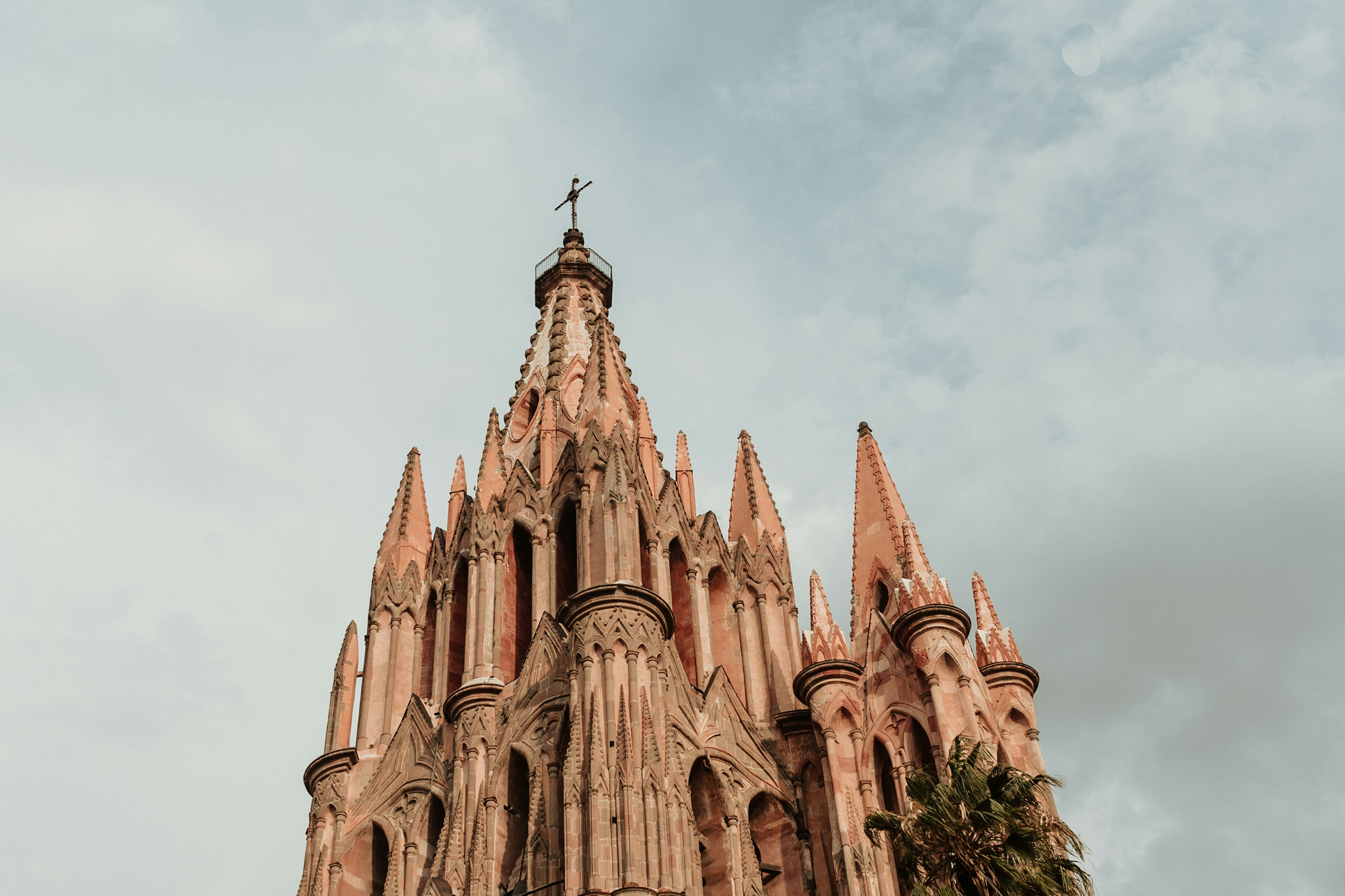 Intricate architectural details of a towering church spire against a cloudy sky. The design features sharp angles and ornate decorations.