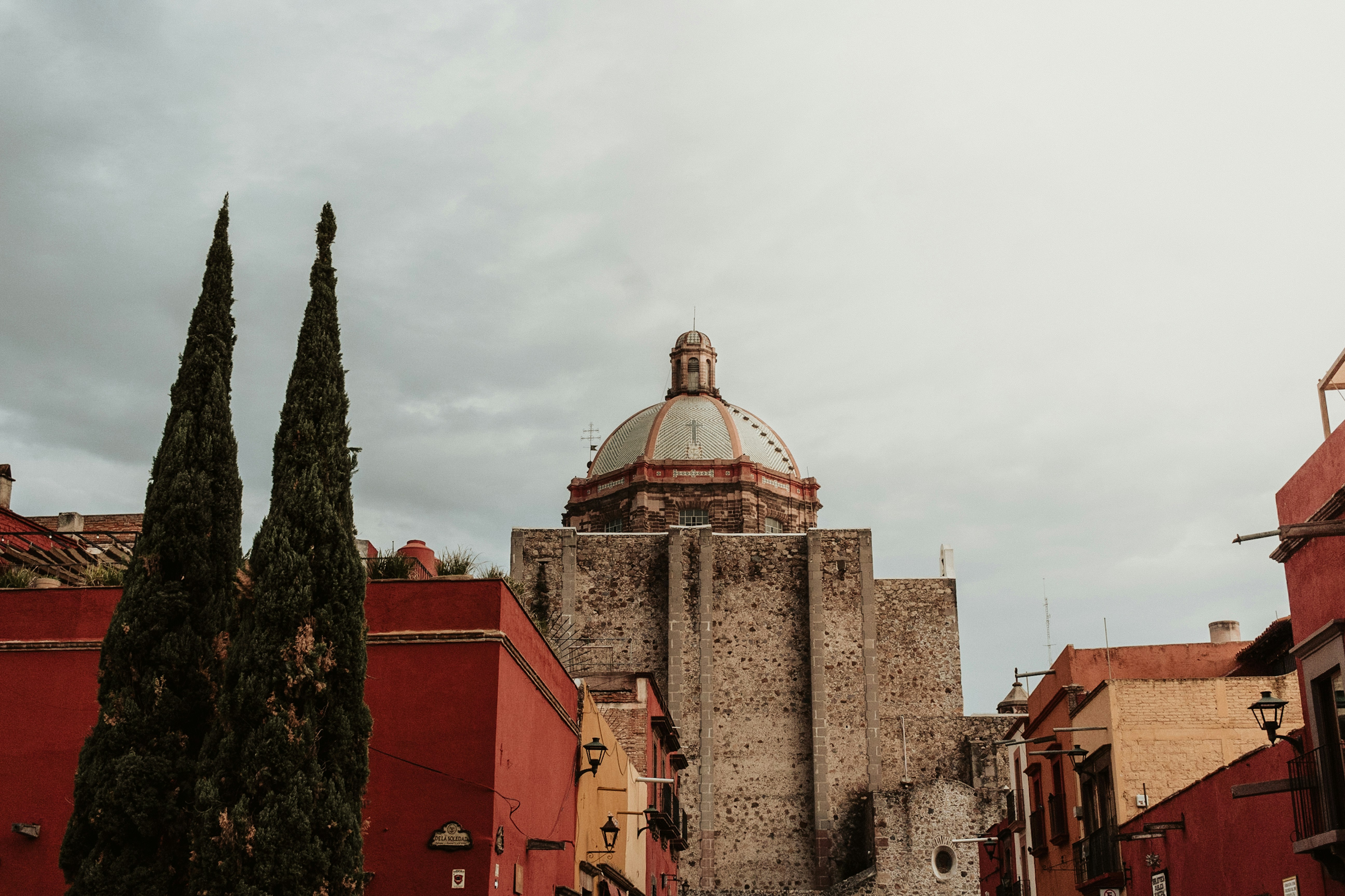 Historic cathedral with a dome surrounded by vibrant red buildings and tall trees under an overcast sky.