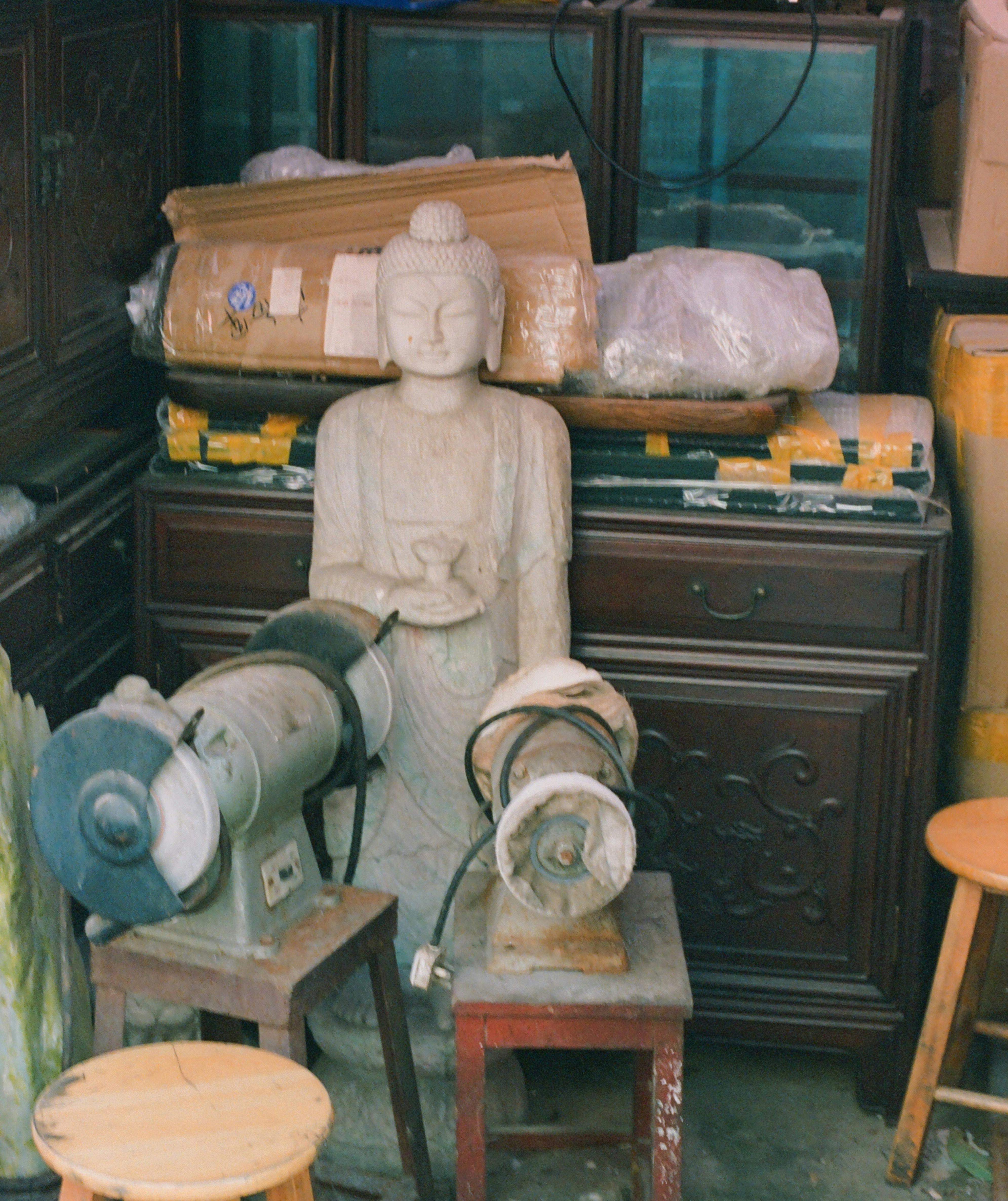 A serene stone statue of a Buddha stands among various tools and boxes in a cluttered workshop. The juxtaposition of tranquility and chaos creates a unique ambiance.