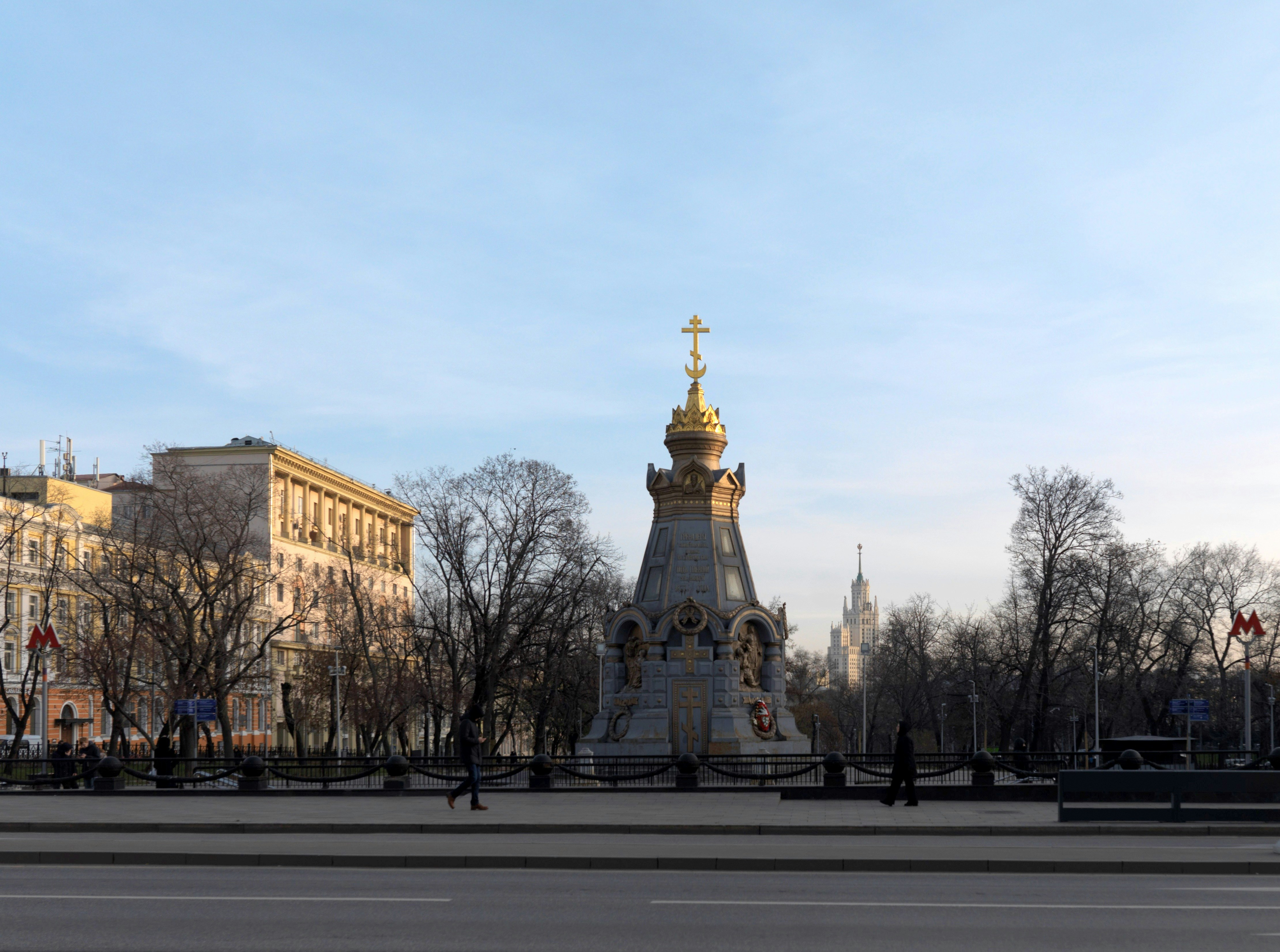 A prominent monument adorned with a golden cross stands in a city square, surrounded by bare trees and historical buildings under a clear sky.