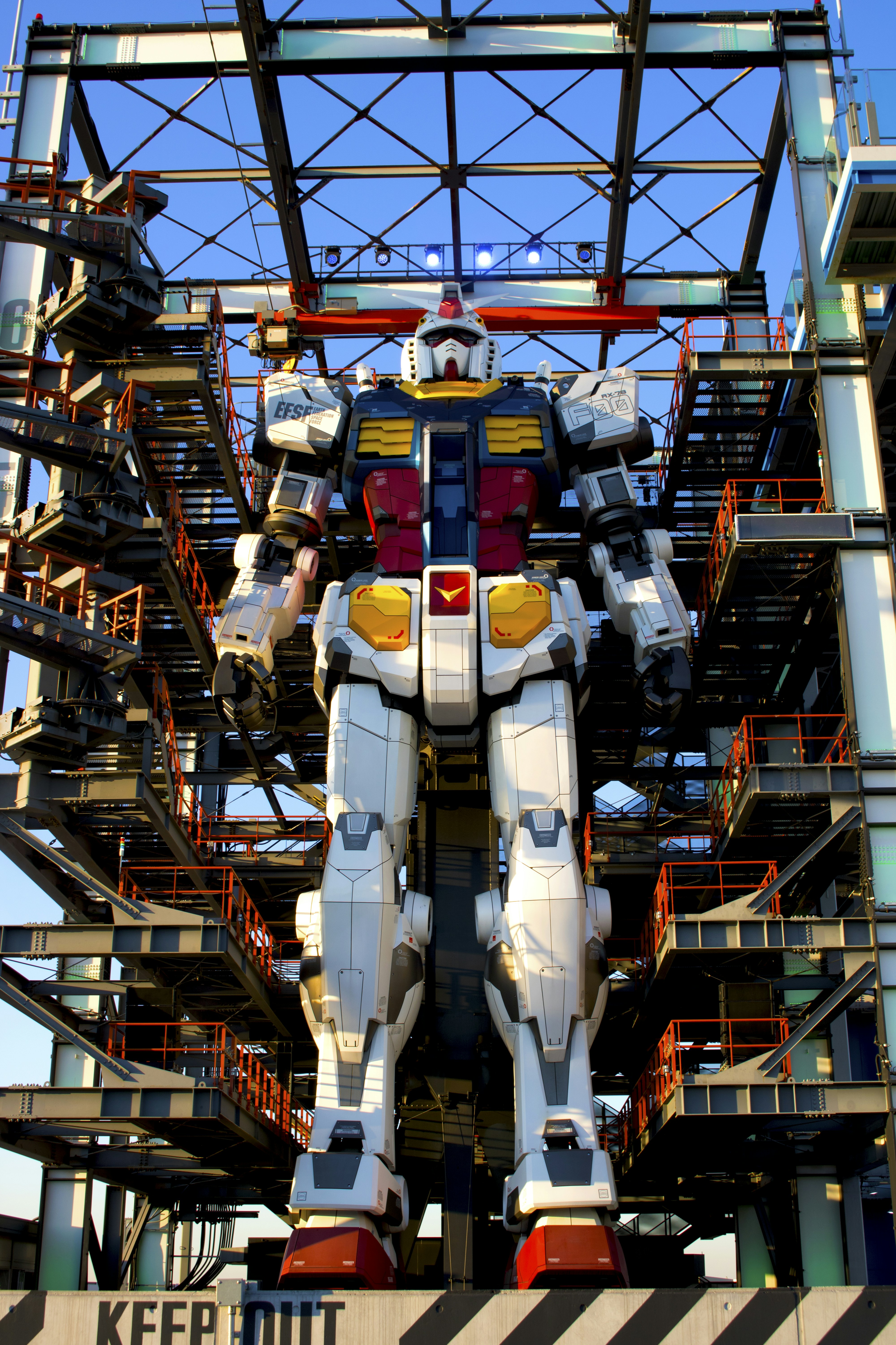 Giant Gundam robot stands tall against a clear blue sky, surrounded by an industrial structure. The scene captures the fusion of technology and artistry.