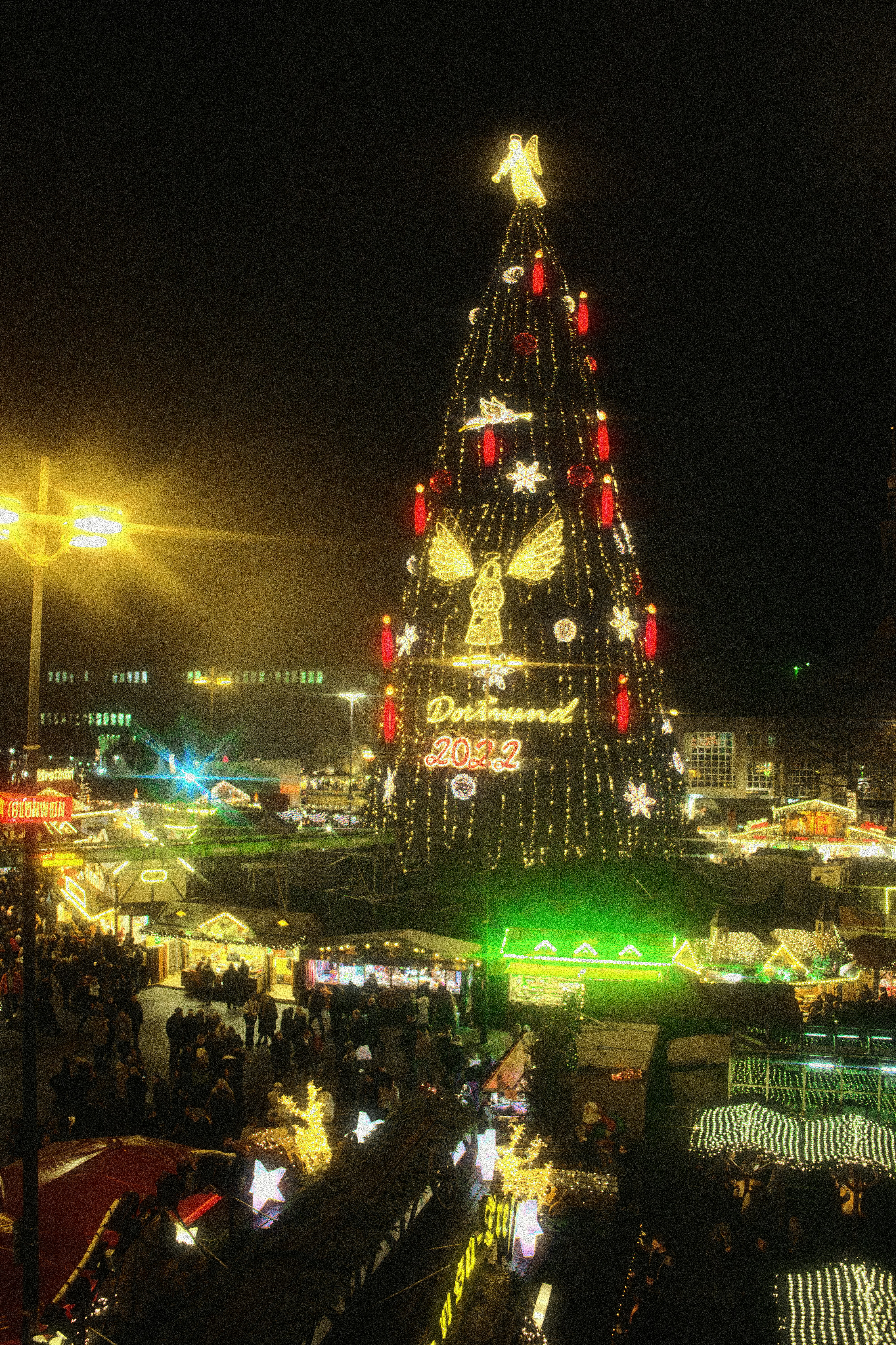 Illuminated Christmas tree towering over a bustling night market adorned with colorful lights and decorations.