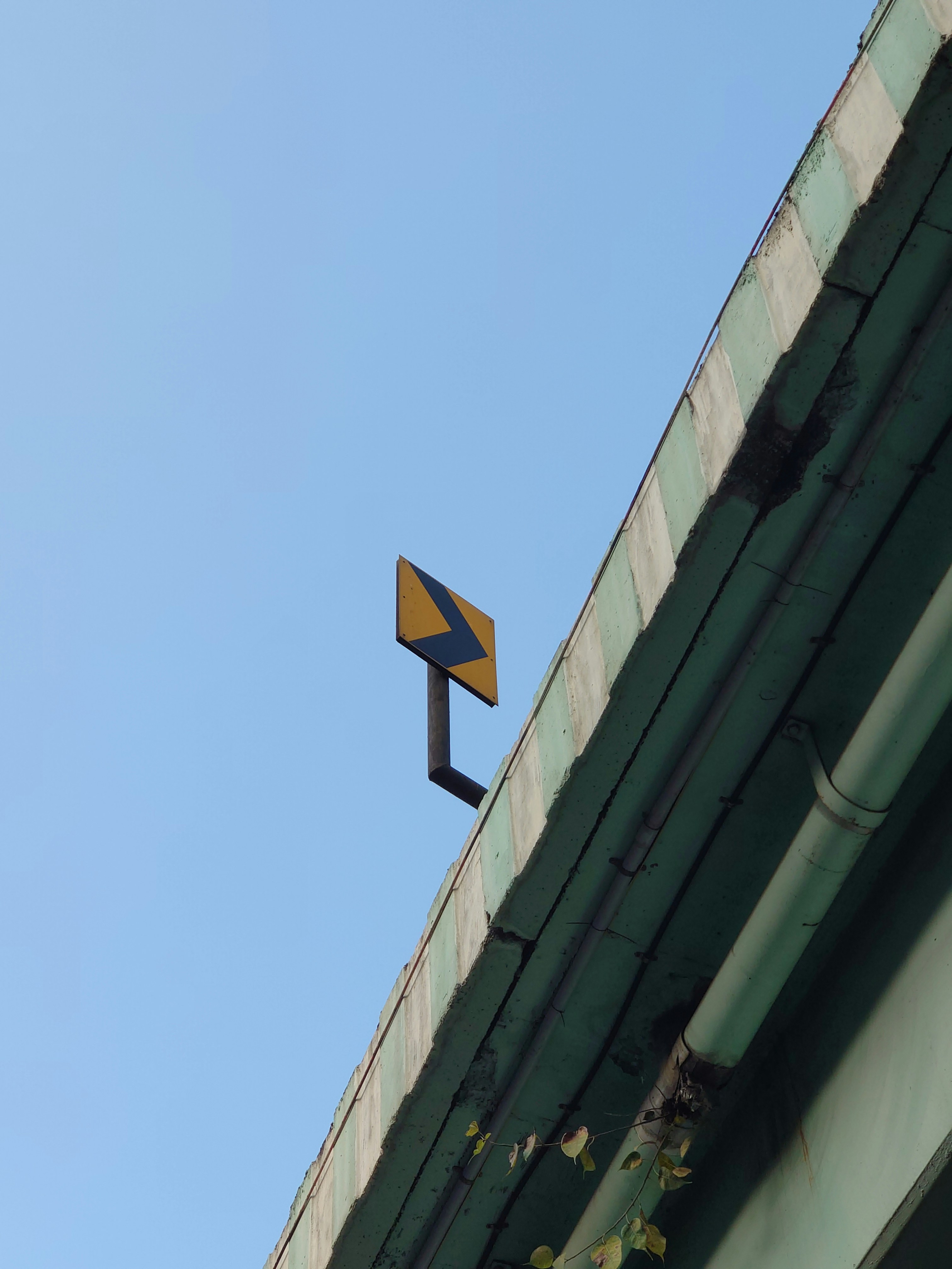 Yellow-and-black chevron road sign on a slim post projects from a green overpass against a clear blue sky.
