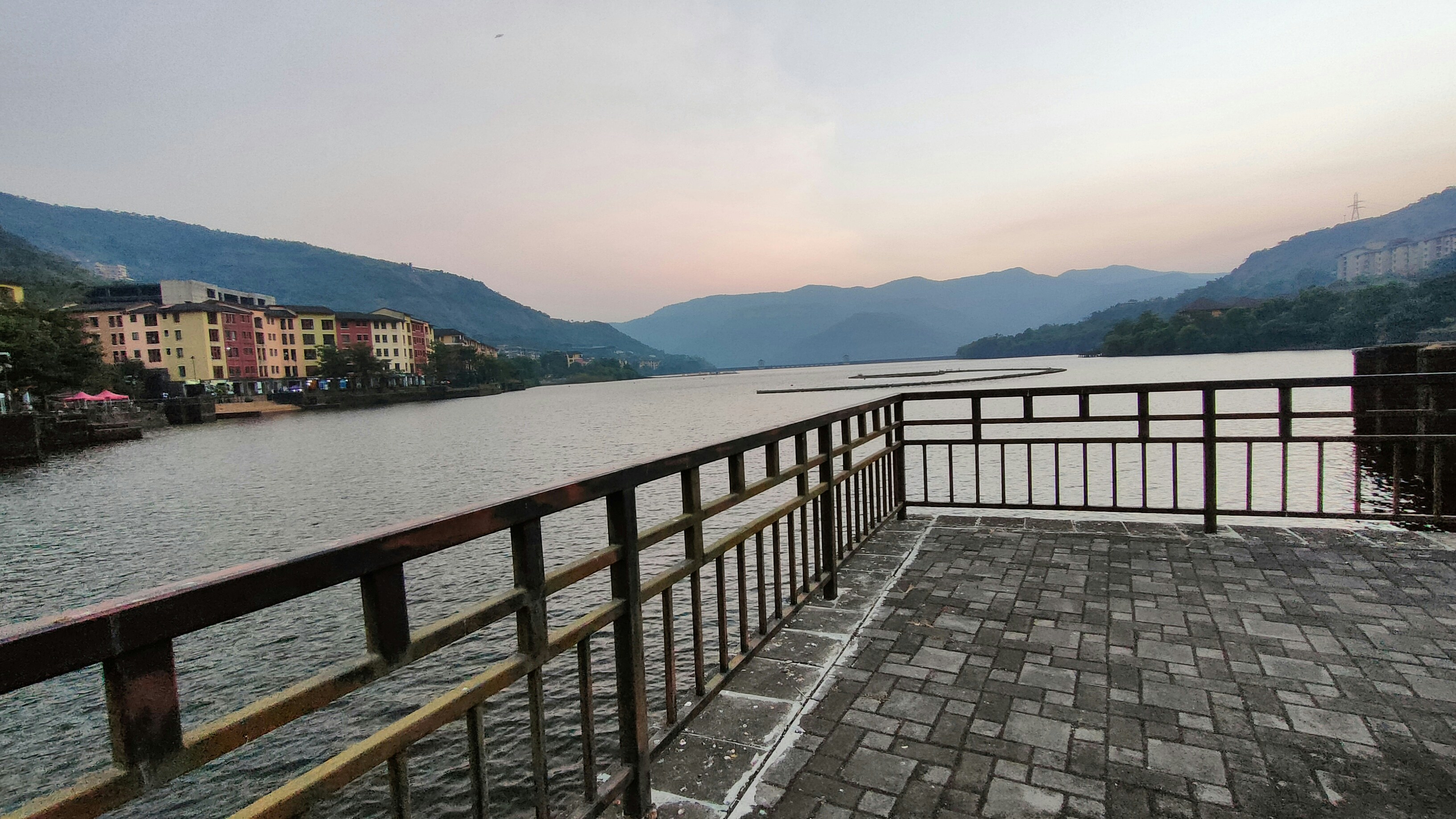 A brick-paved riverside promenade with a wooden railing along calm water. Pastel-lit buildings line the left shore as hazy mountains rise in the distance.