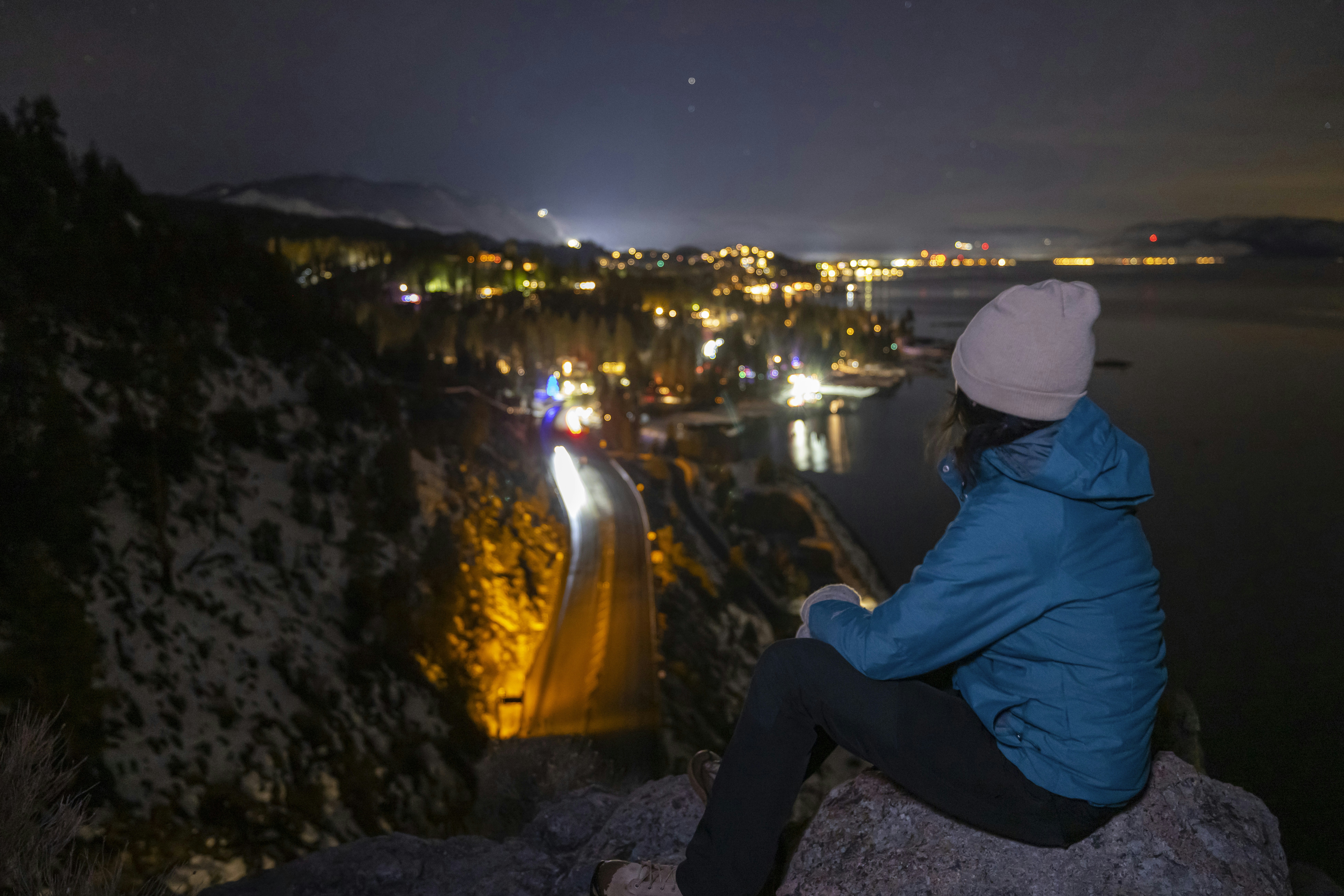 Person in a blue jacket and white beanie gazing at city lights from a rocky vantage point at night.