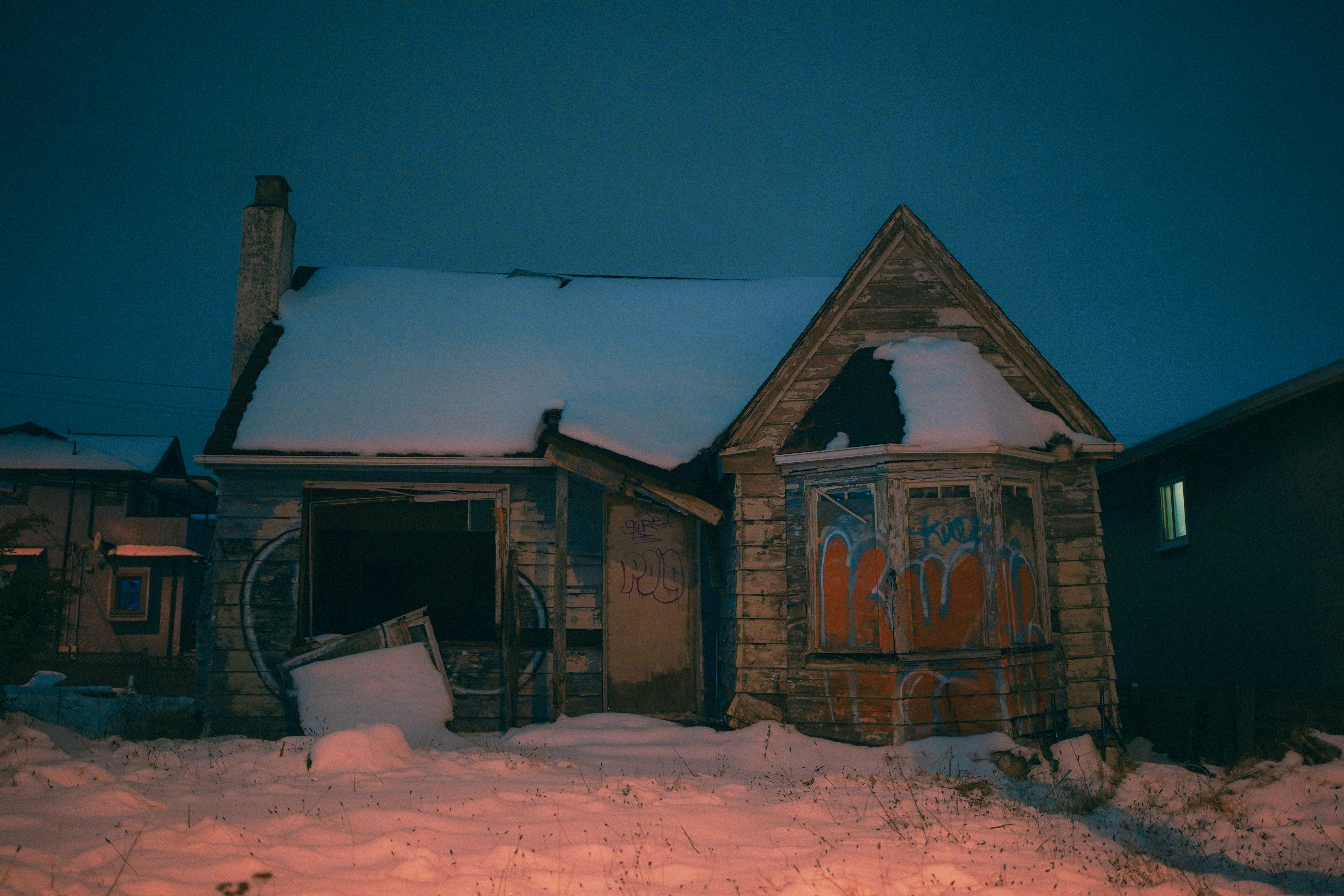 Abandoned house with graffiti, partially covered in snow, under a twilight sky.