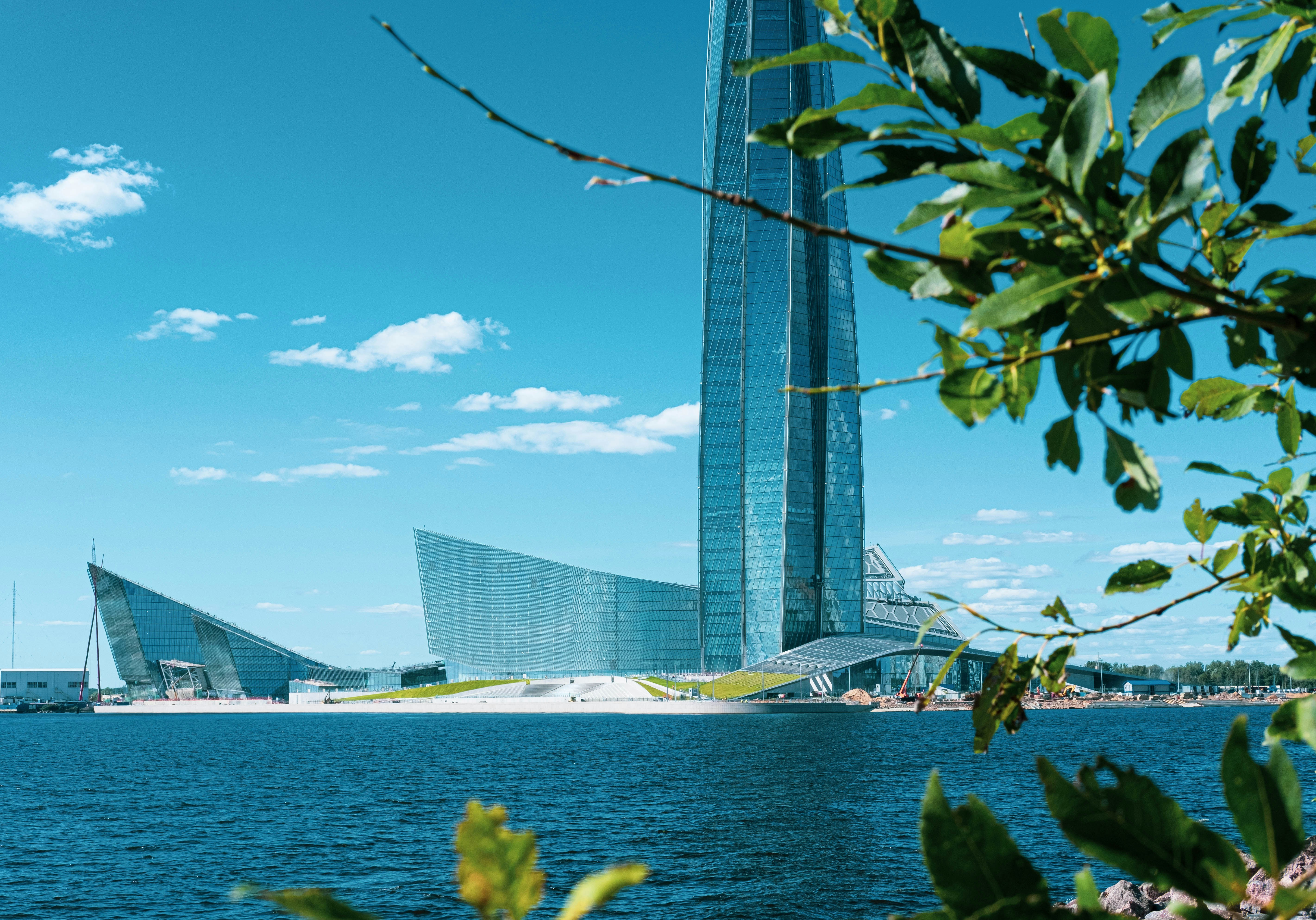 Modern skyscraper rises beside a tranquil body of water, framed by green foliage under a clear blue sky.