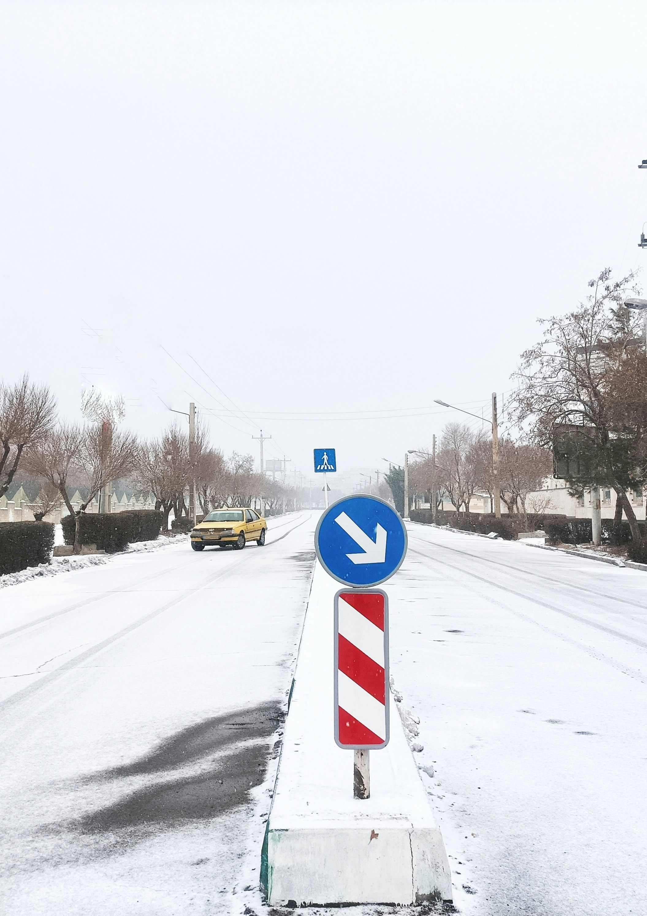 Snow-covered street with a blue circular arrow sign on a concrete barrier in the foreground and a yellow car in the distance.