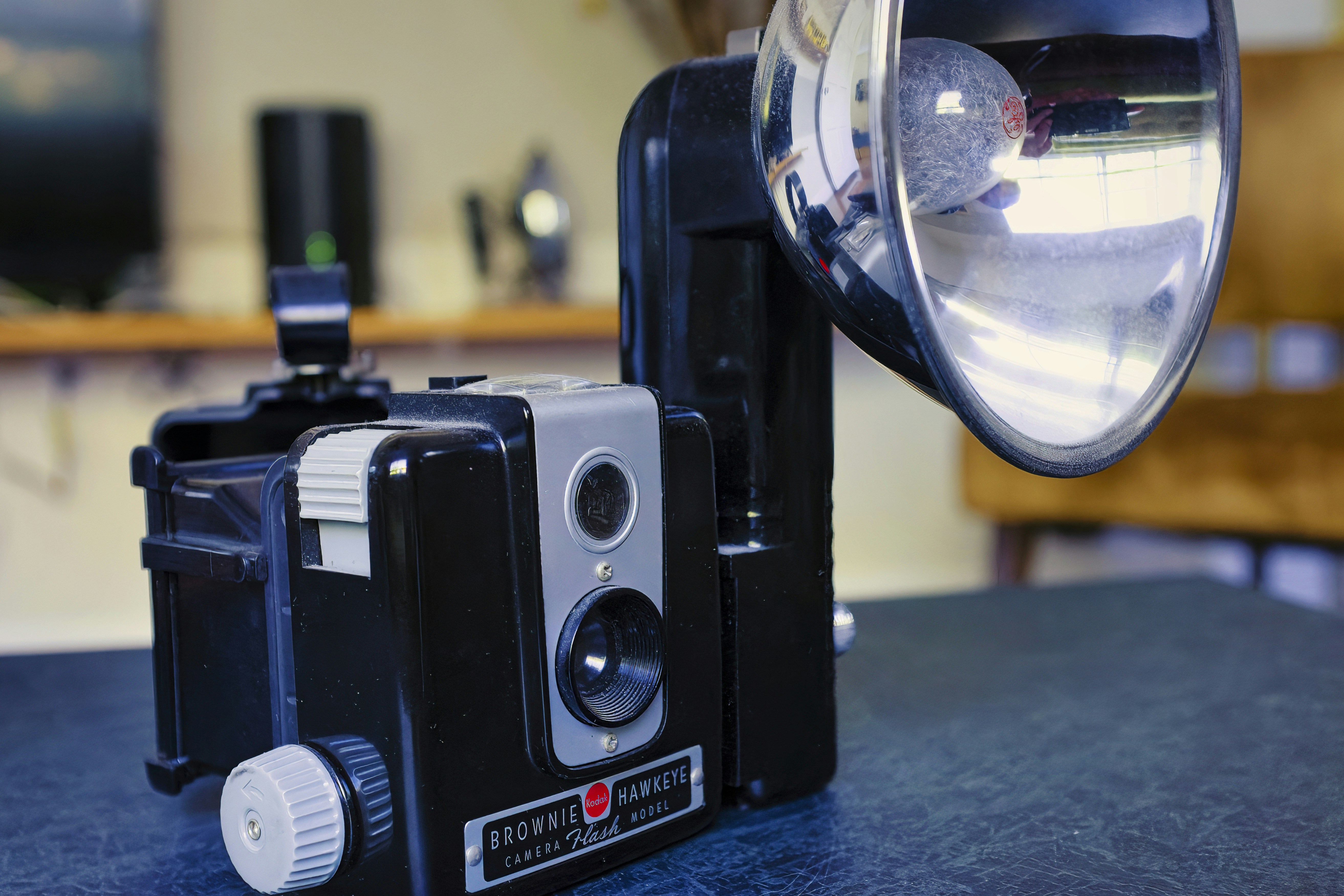 Vintage Brownie Hawkeye Flash Model Camera With External Flash Unit on Blue Tabletop - A vintage Brownie Hawkeye Flash Model camera is positioned on a blue tabletop. The camera has an external flash unit attached to the top, and the camera's lens and flash reflector are both in focus. The image captures the camera from a slightly elevated perspective, highlighting its classic design and features.