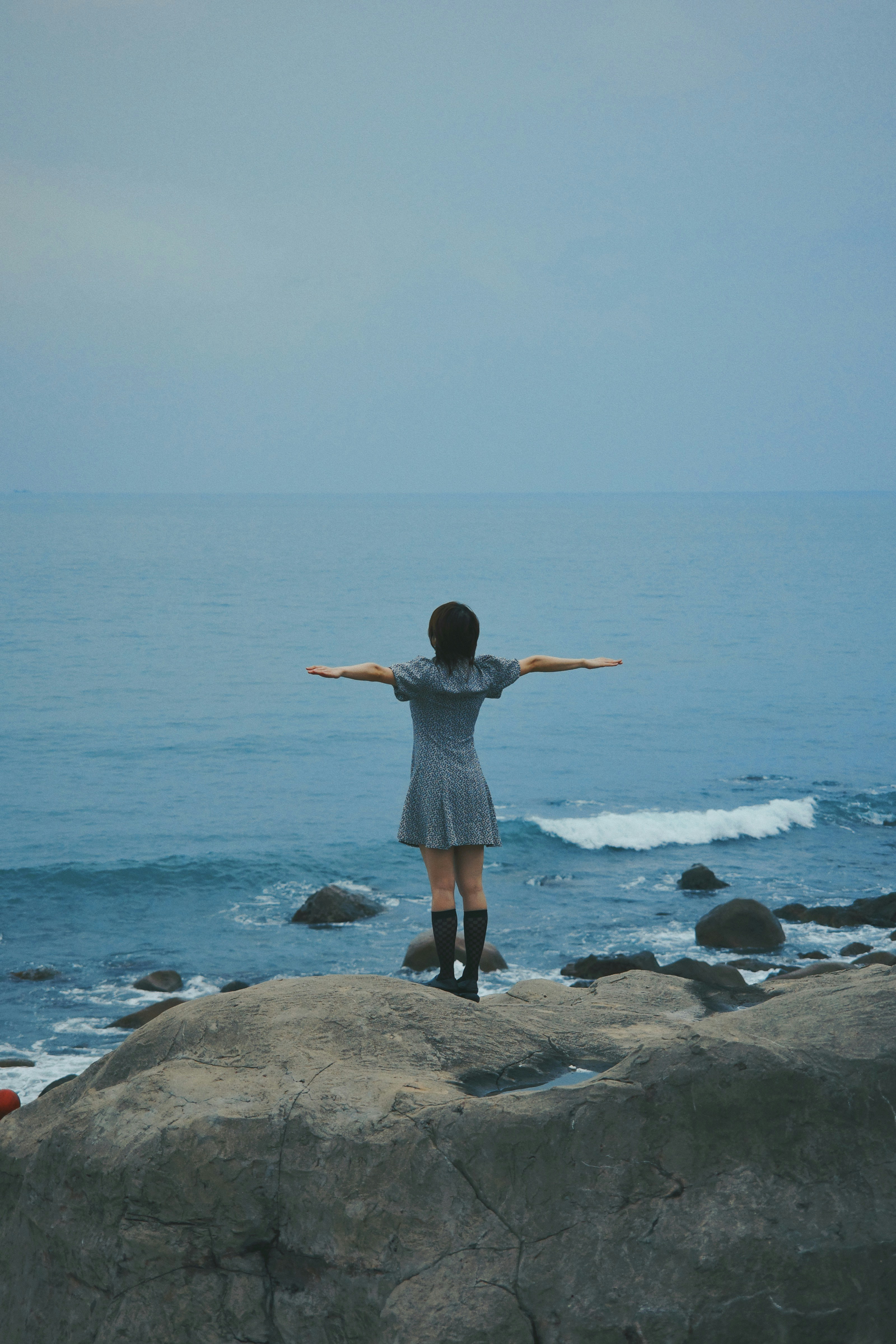 Individual standing on rocky shore with arms outstretched, gazing at the calm sea under a cloudy sky.