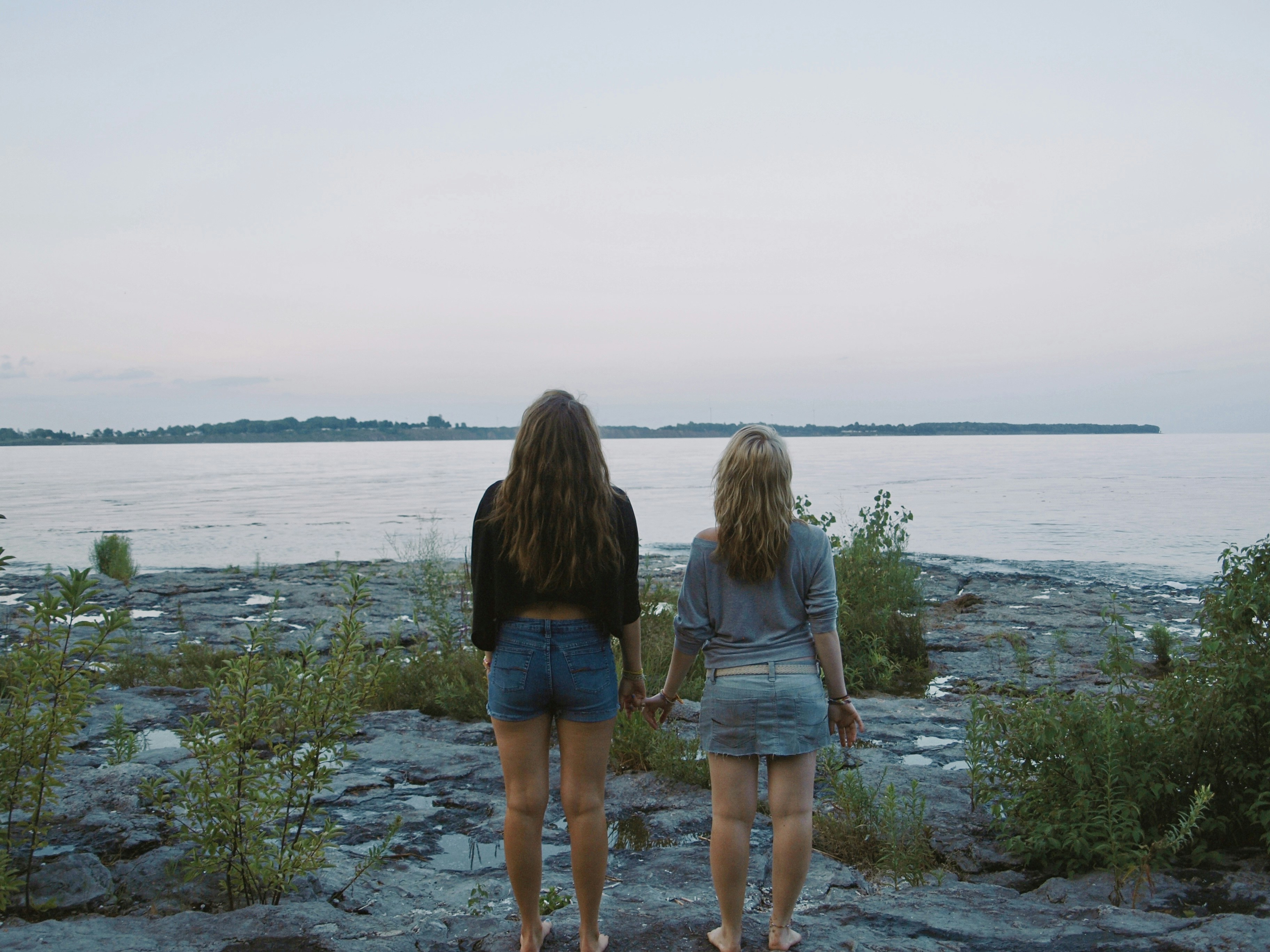 Two people stand on rocky shoreline overlooking a calm lake under a pastel sky.